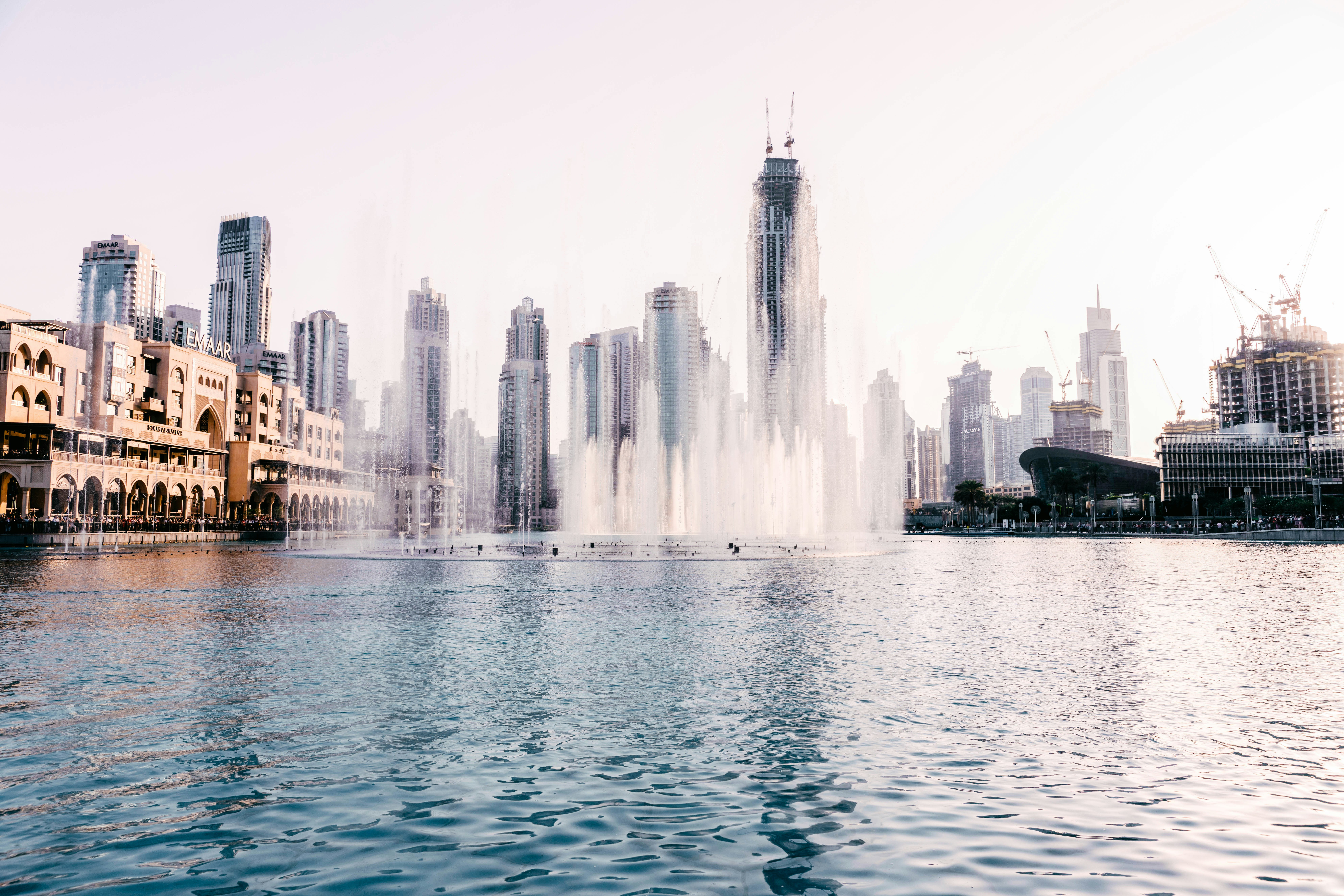 Waterfront fountain jets rise against a pastel-dawn skyline, with calm water reflecting the towers.