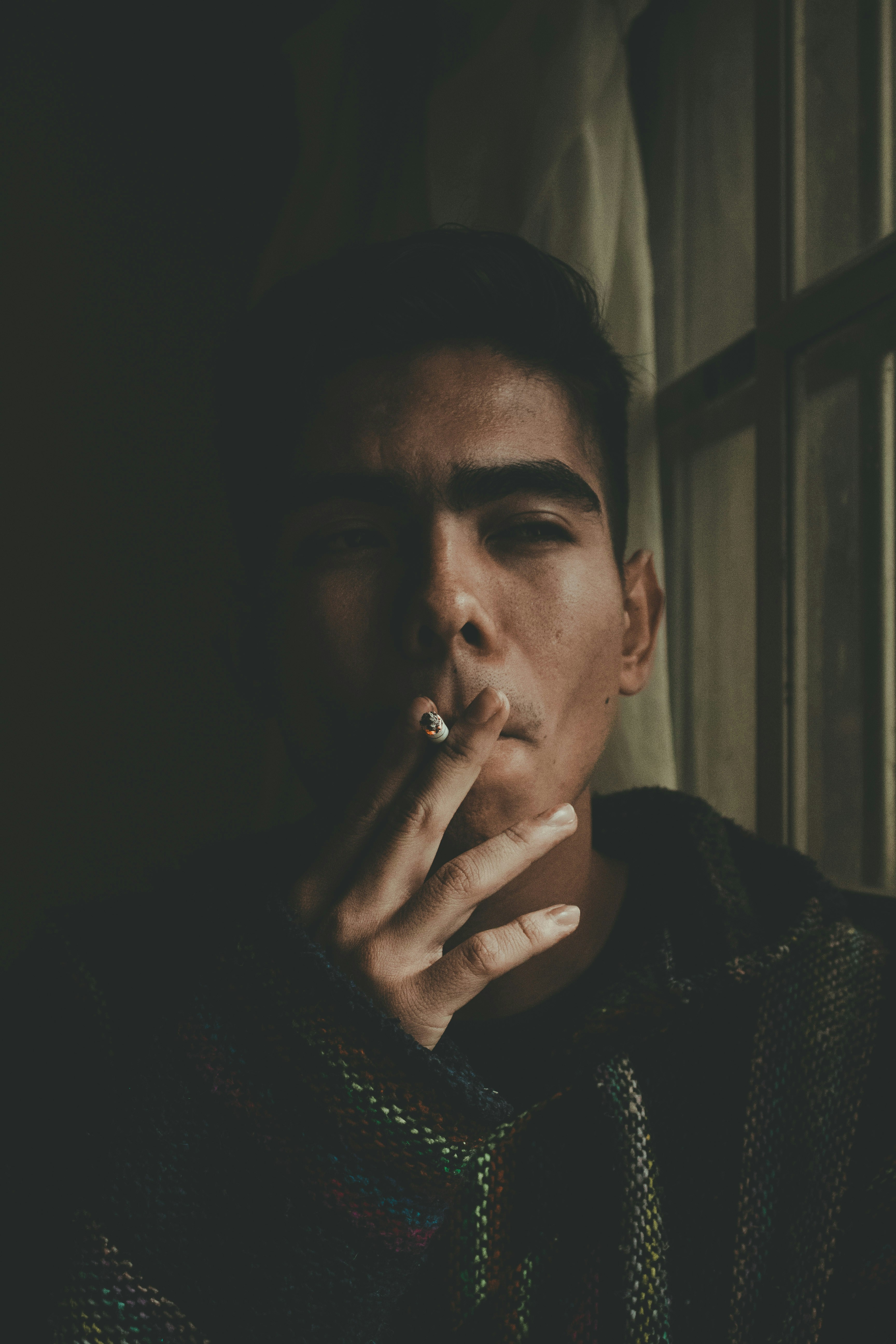 Young man exhaling smoke while standing near a window, enveloped in dim lighting. The atmosphere evokes a sense of contemplation and introspection.