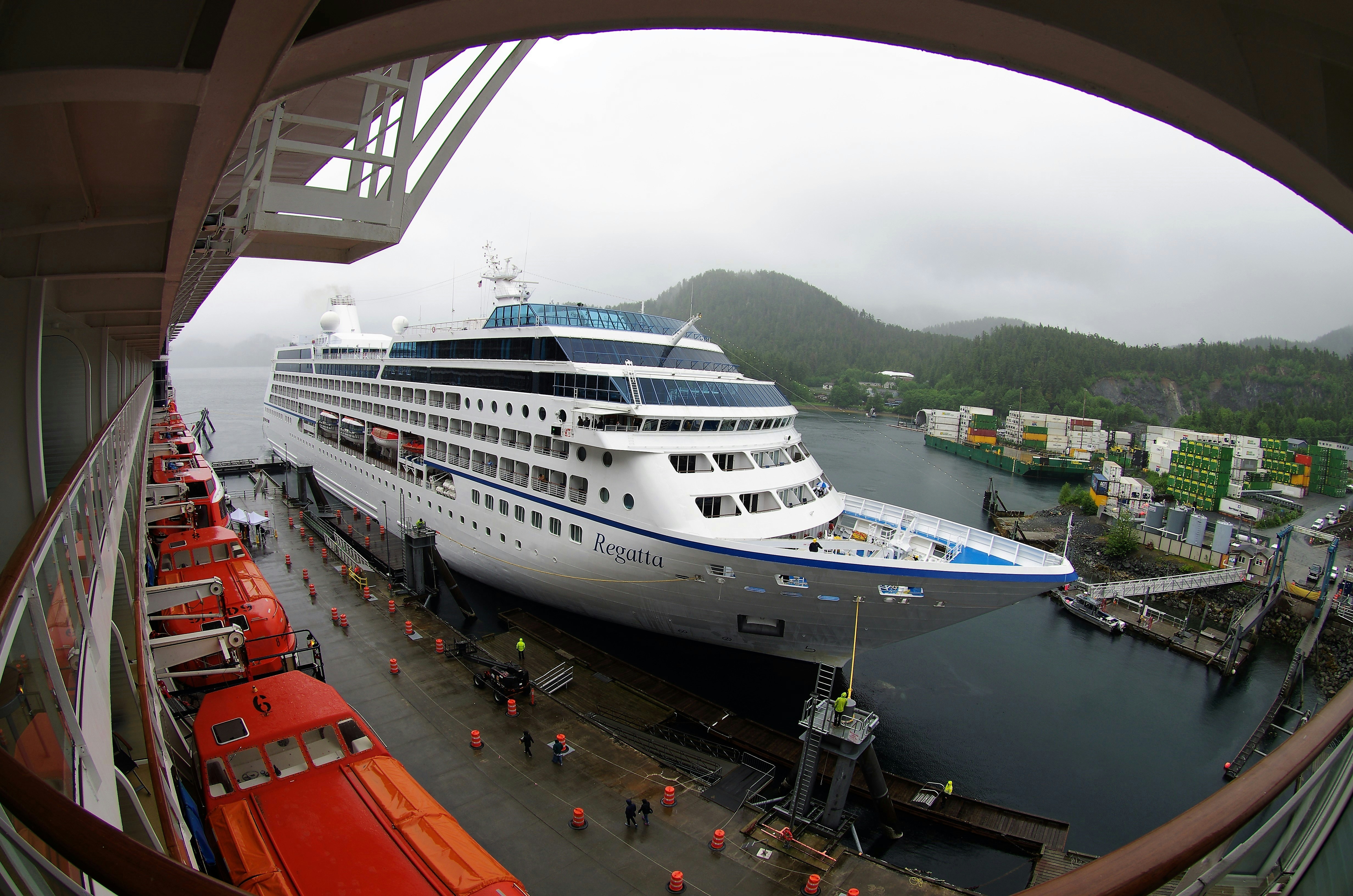 Large cruise ship docked at a harbor, surrounded by mountainous terrain and colorful containers. The scene captures a blend of maritime activity and natural beauty.
