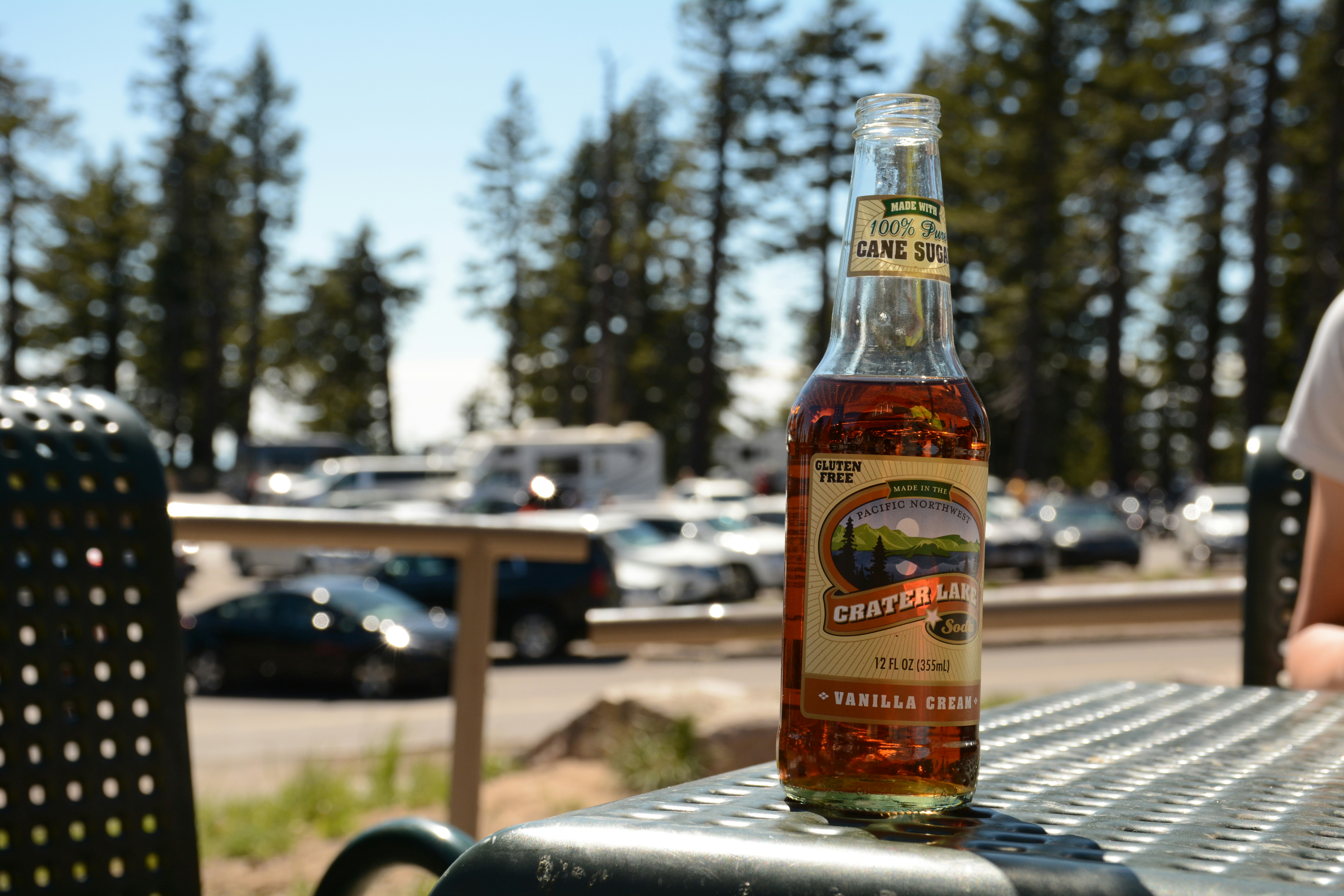 Crater Lake Vanilla Cream soda bottle rests on a table, surrounded by a lush forest backdrop and parked vehicles. The scene captures a moment of relaxation in a vibrant outdoor setting.