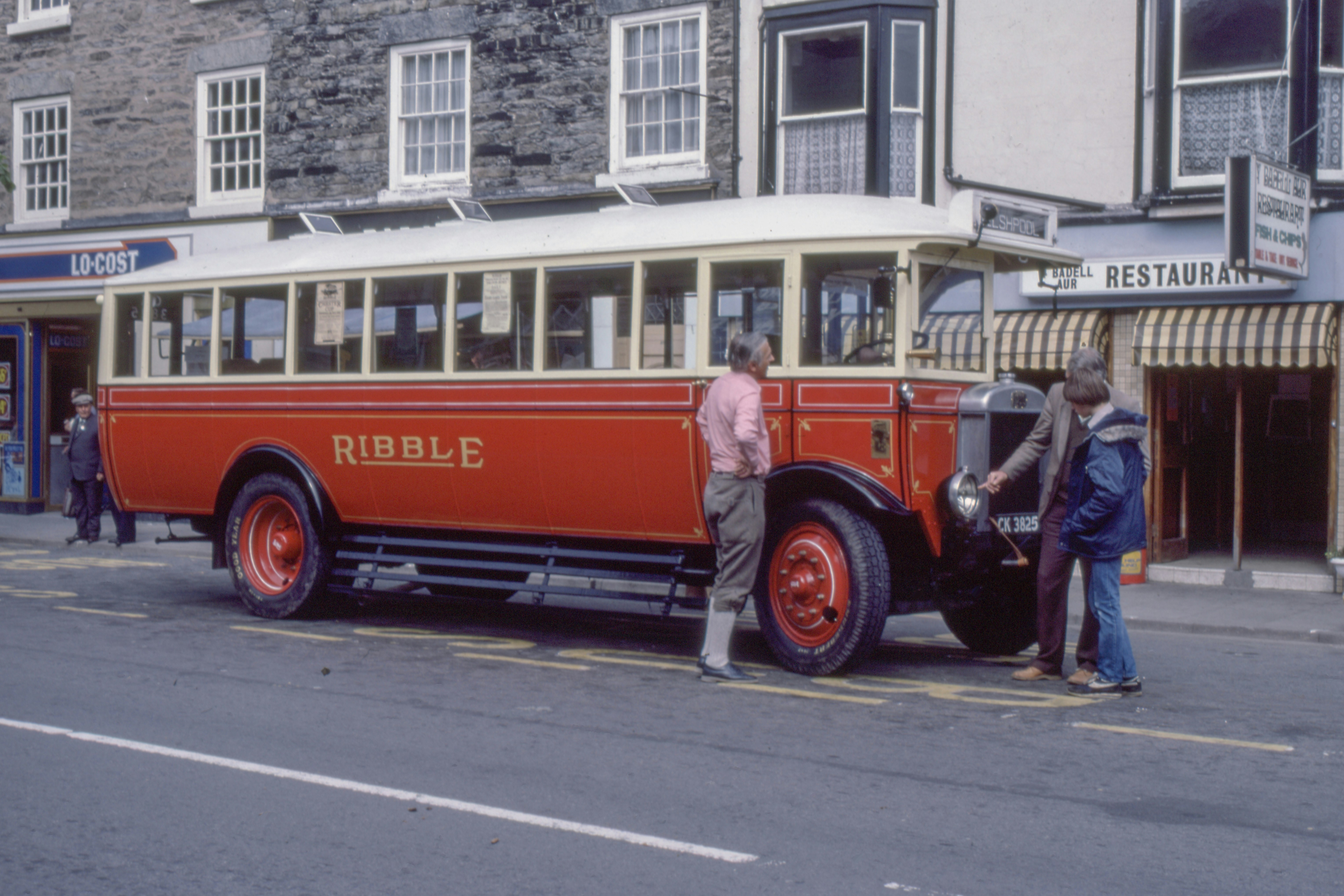 Classic red Ribble bus parked on a quaint street, with two men engaged in conversation beside it. The scene reflects a nostalgic era of transportation.