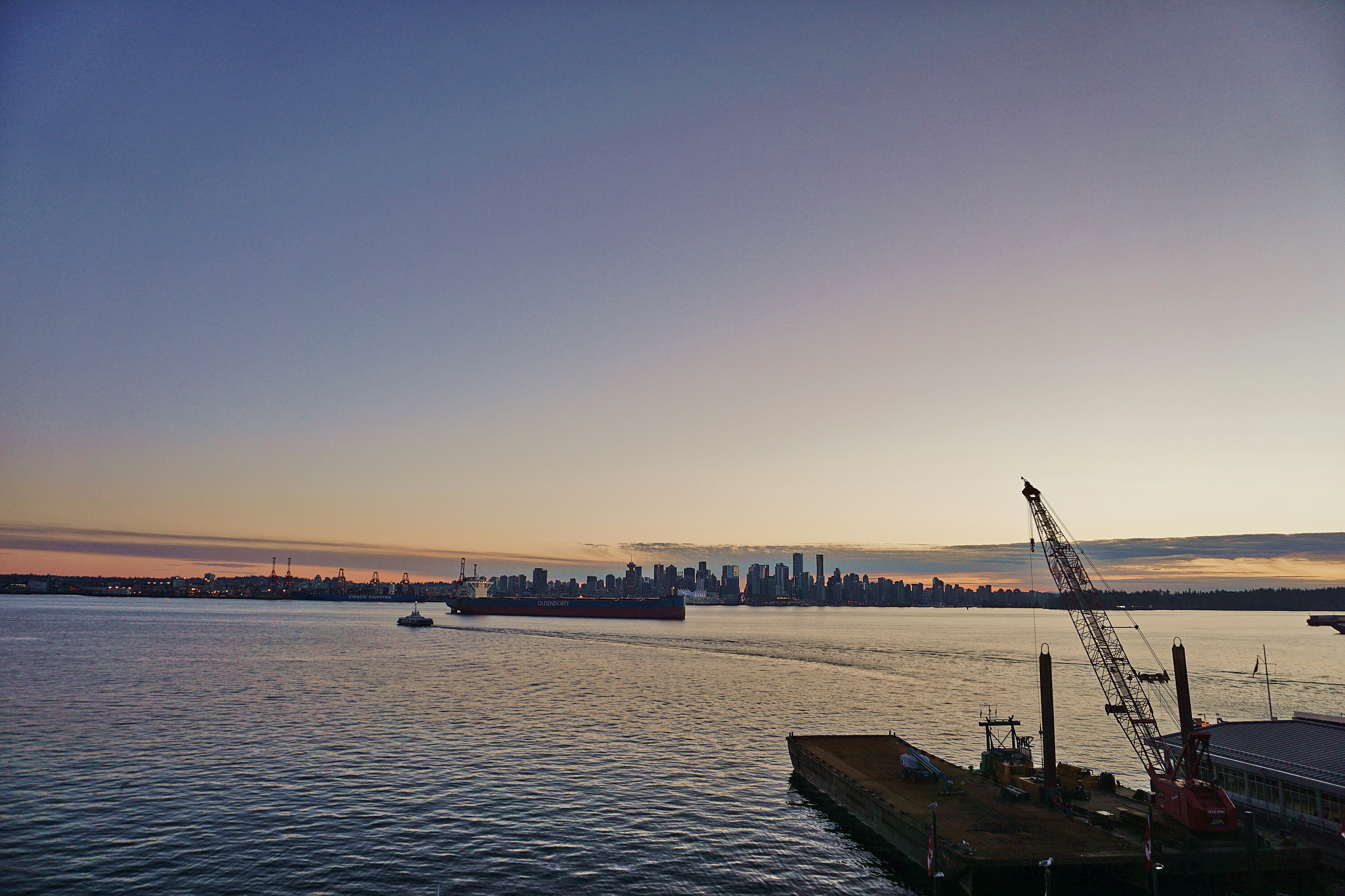 Cargo ship gliding across calm waters towards a city skyline at dawn, with a dock and crane in the foreground.