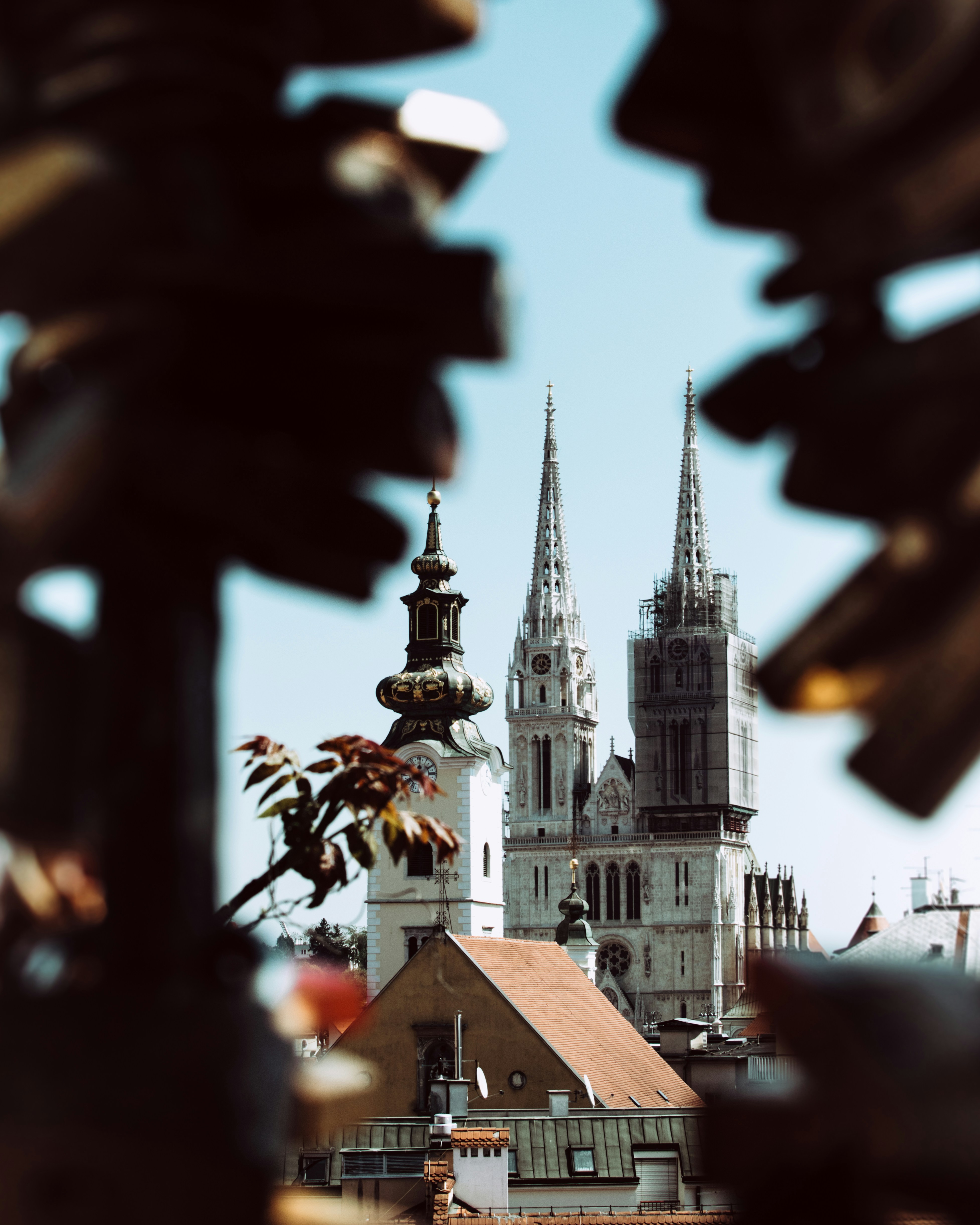 Two towering spires of a historic cathedral rise majestically above rooftops, framed by blurred foliage in the foreground.