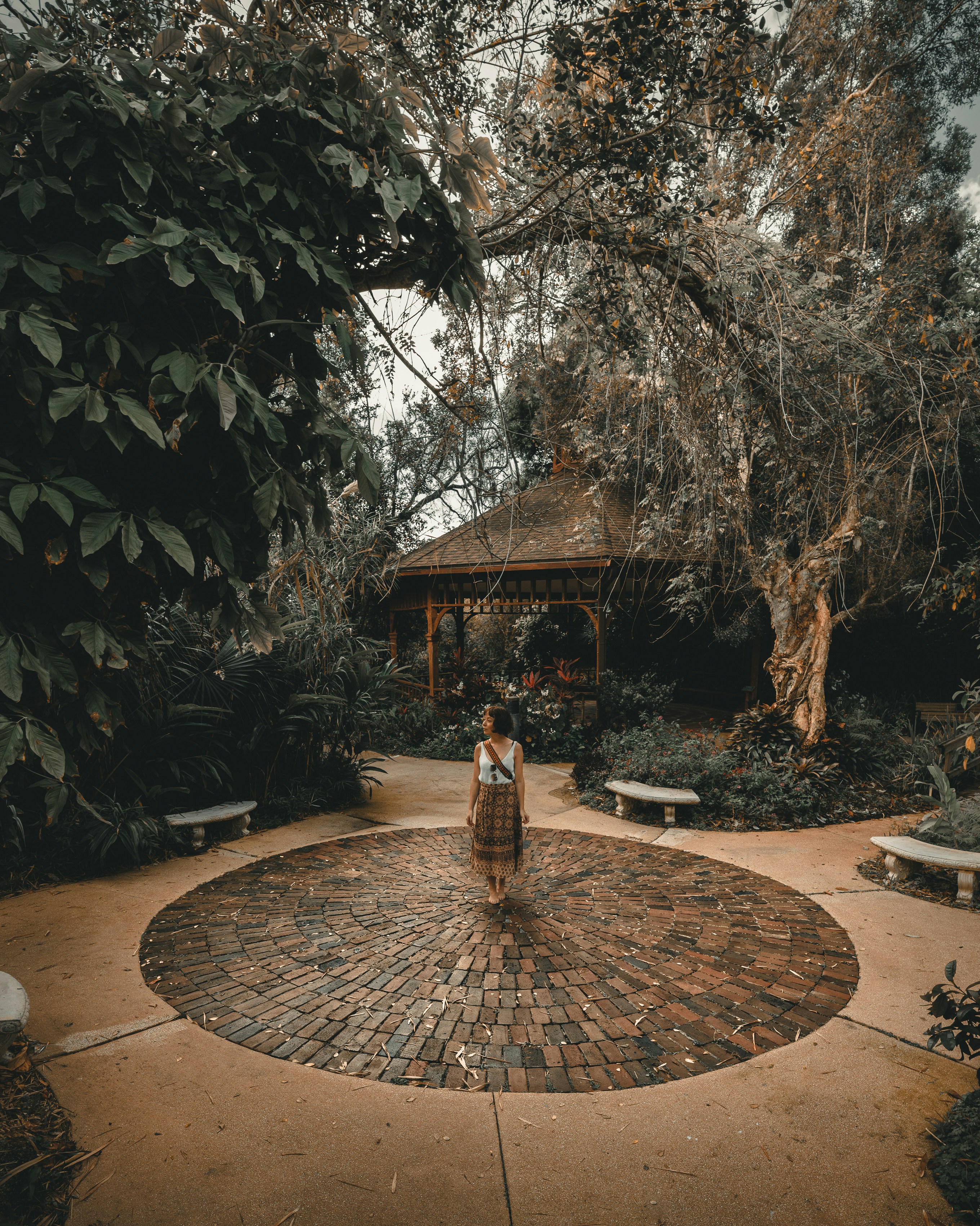 A solitary figure walks along a circular stone path in a serene garden, surrounded by lush greenery and a rustic gazebo. The atmosphere evokes tranquility and reflection.