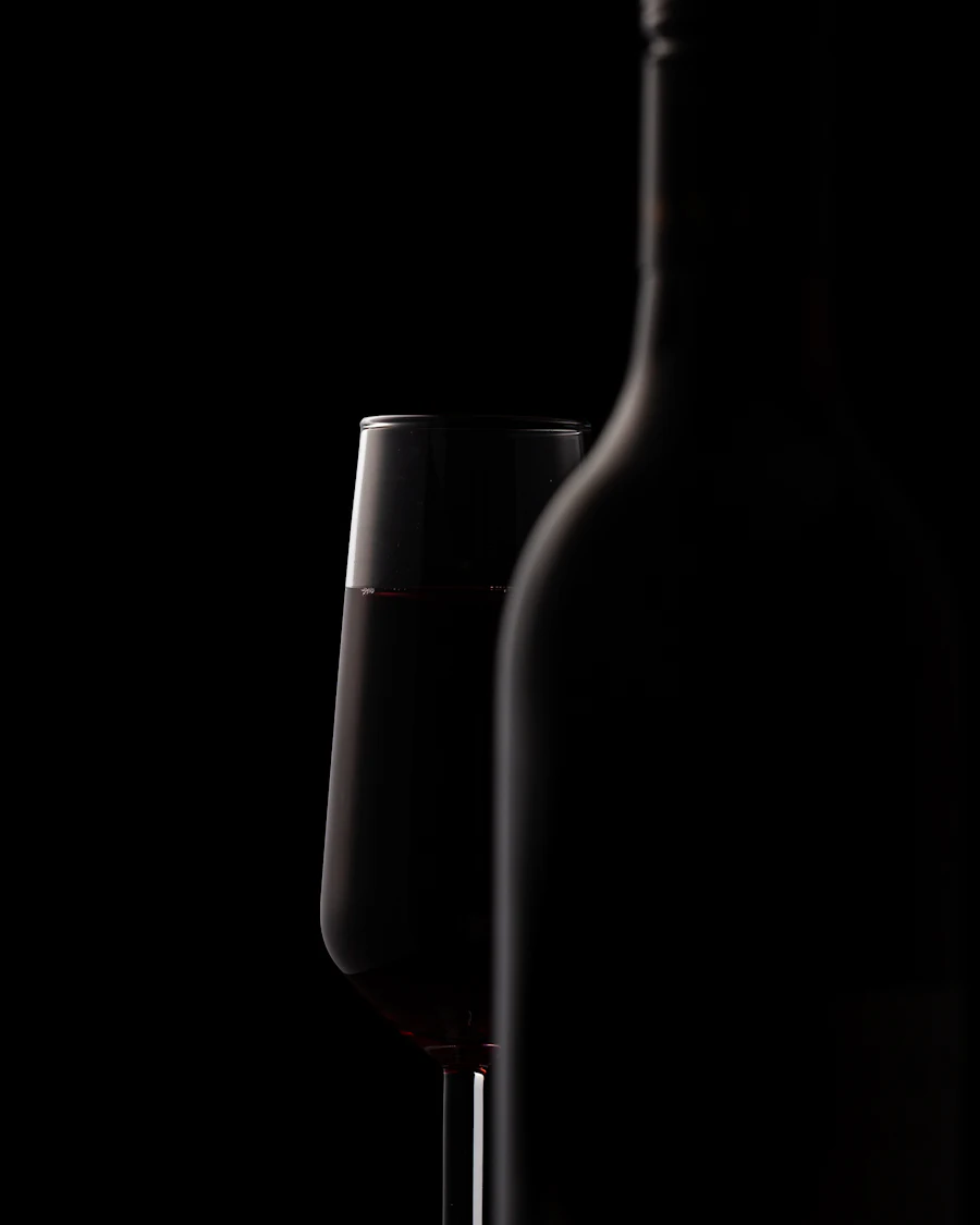 A dark, moody still life with a wine glass partially filled with red wine, seen behind the blurred silhouette of a bottle. The scene is lit softly against a black background.