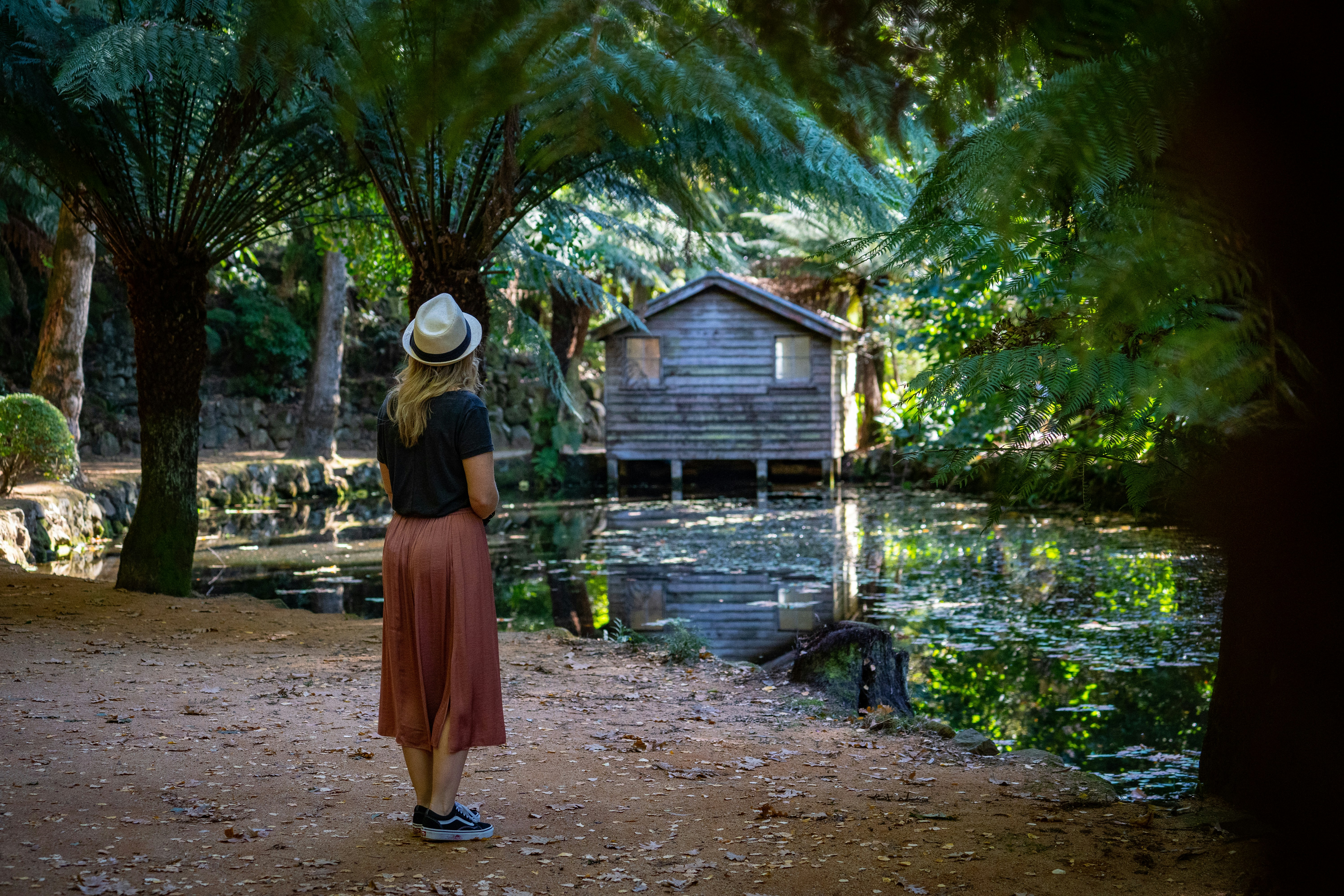 A woman in a hat stands contemplatively by a tranquil pond surrounded by lush greenery and a rustic cabin. Soft light filters through the trees.