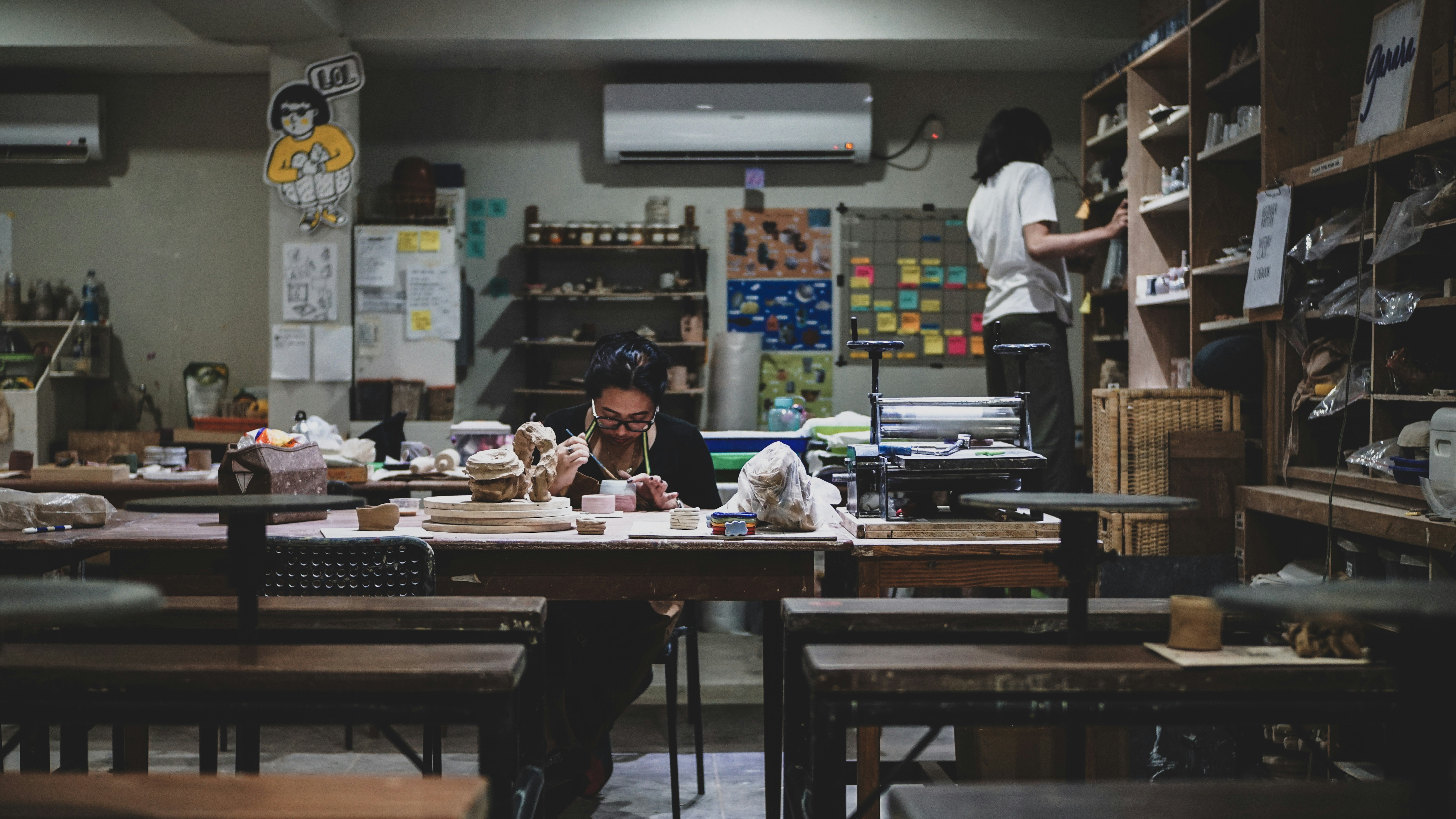 Person engaged in pottery work at a cluttered workshop table, with shelves filled with materials and supplies in the background.