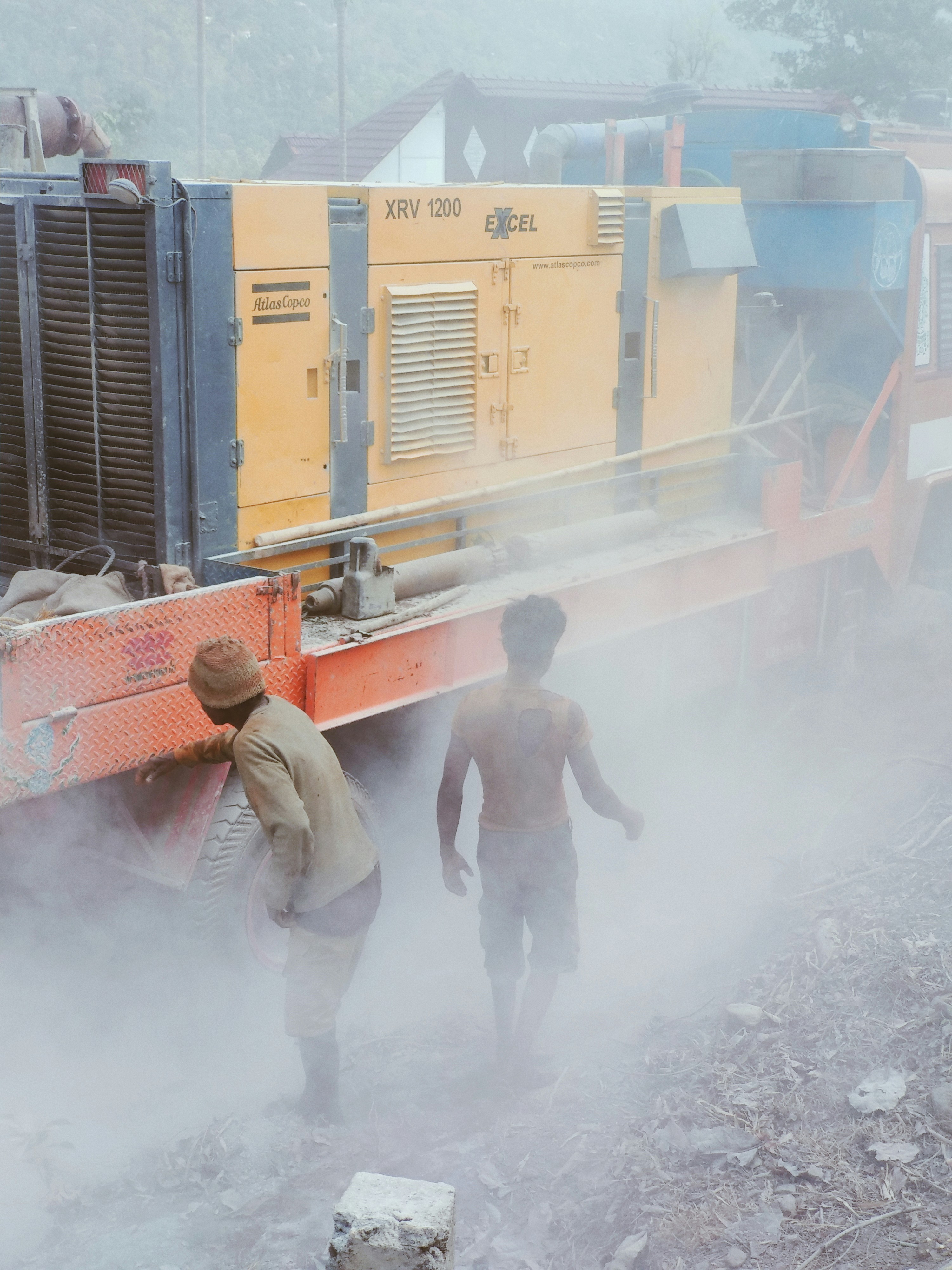 Two workers engaged in manual labor near a large industrial machine, enveloped in a cloud of dust. The scene captures the essence of hard work in challenging conditions.