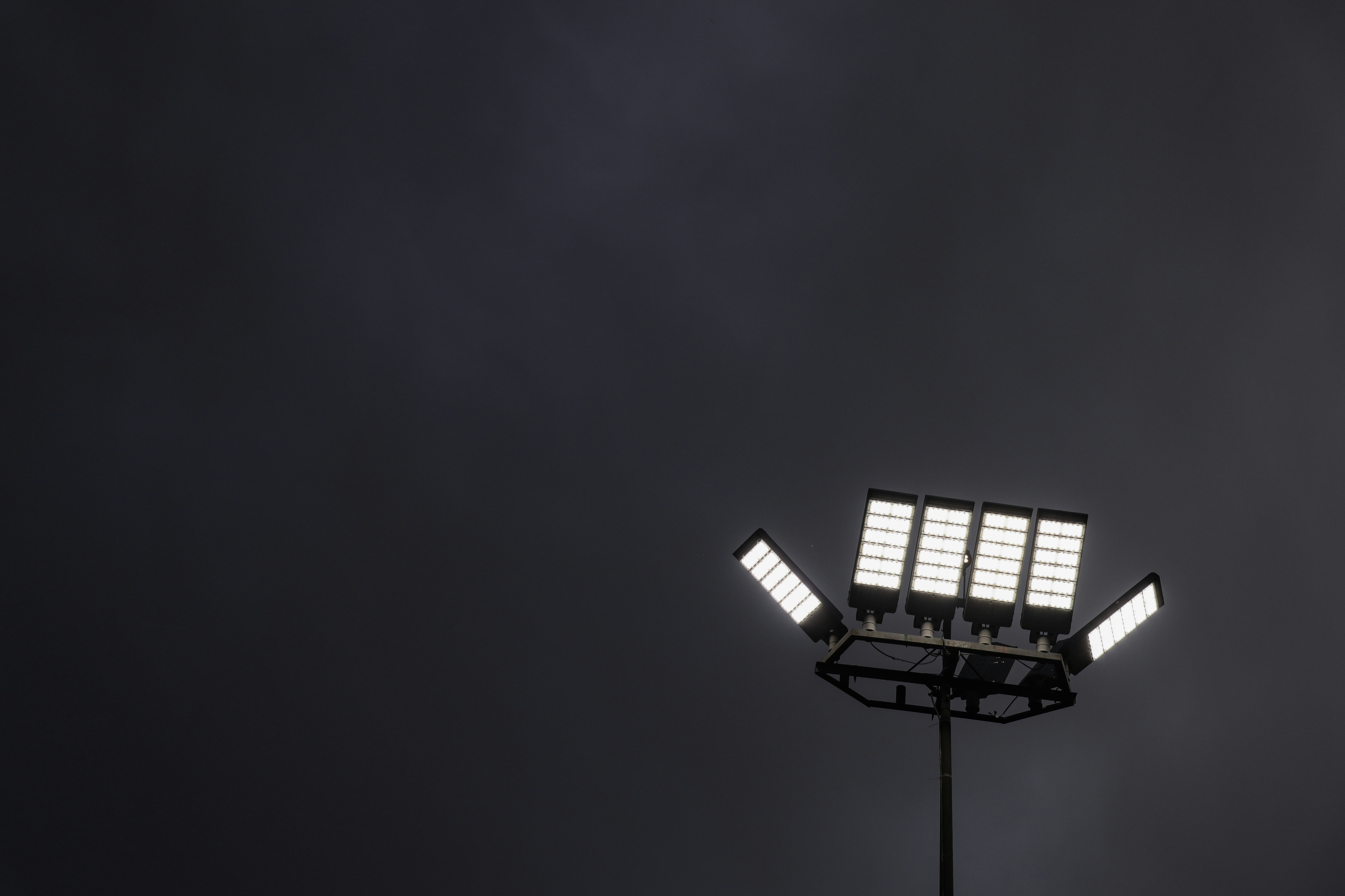 High-powered floodlight standing tall against a dark, overcast sky, casting a stark contrast. The lights illuminate the surrounding area, creating a focal point in the shadowy ambience.