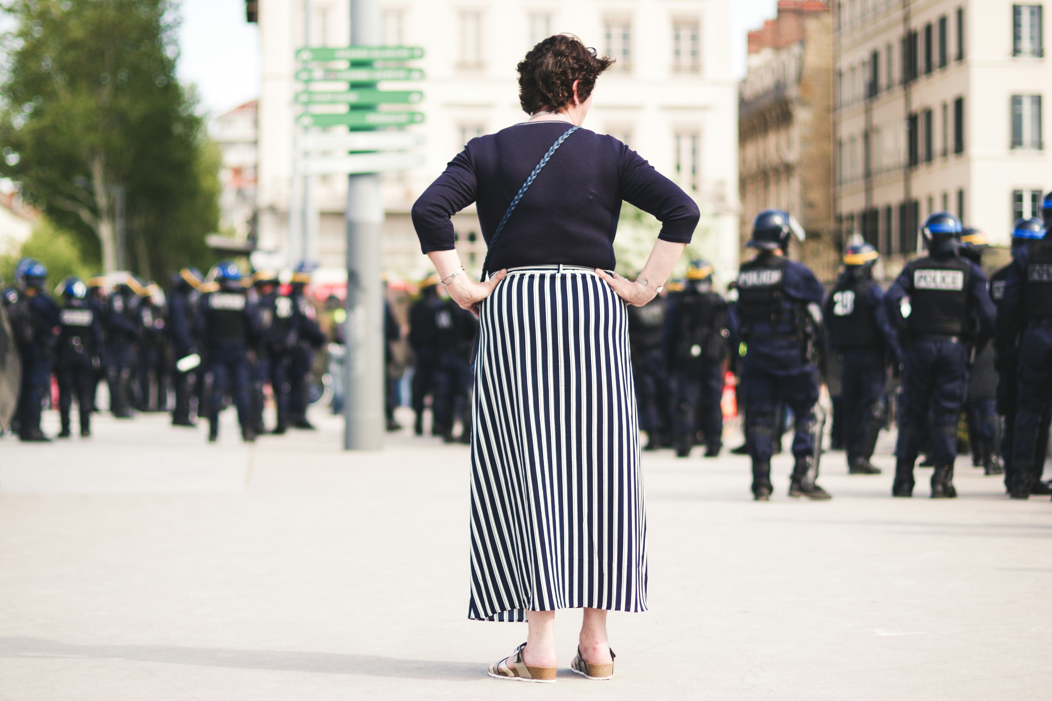 Woman in a striped skirt stands with hands on hips, facing a line of police officers in riot gear. The scene captures a moment of tension and defiance.