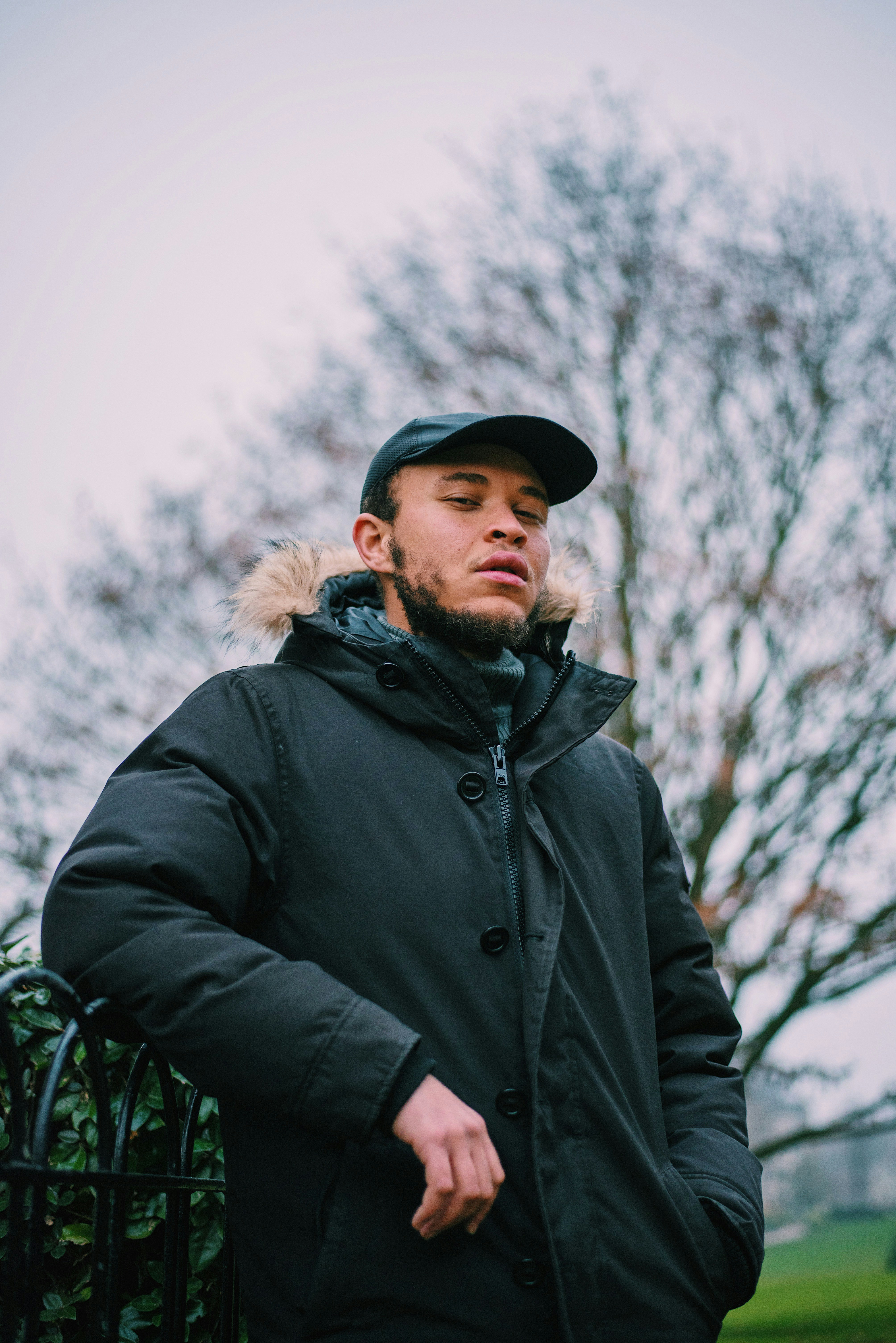 A young man in a black parka leans against a wrought-iron fence, surrounded by a muted winter landscape. His contemplative expression contrasts with the soft, overcast sky.