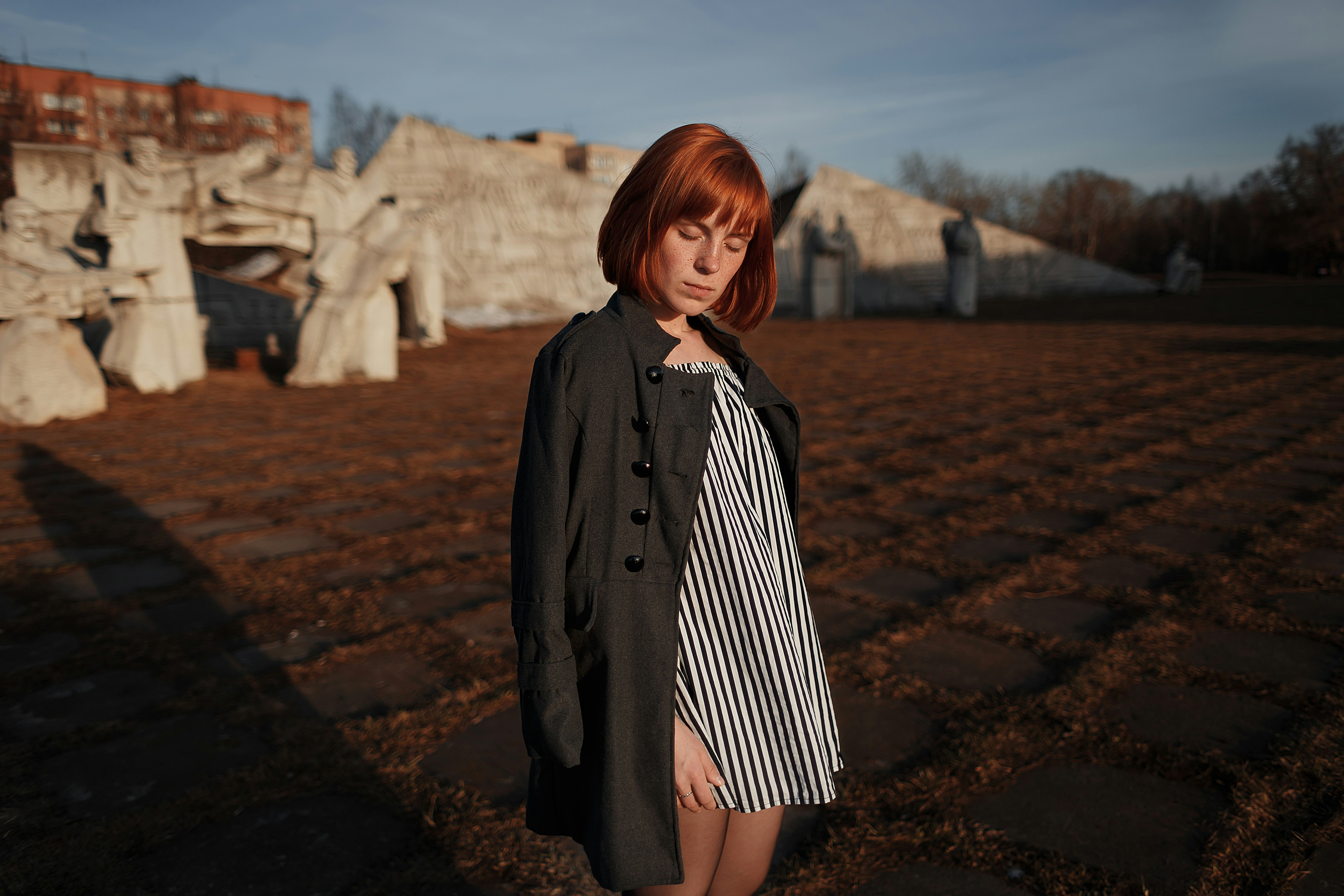 Model in a striped dress and dark coat stands thoughtfully in a sunlit outdoor setting with abstract sculptures in the background.