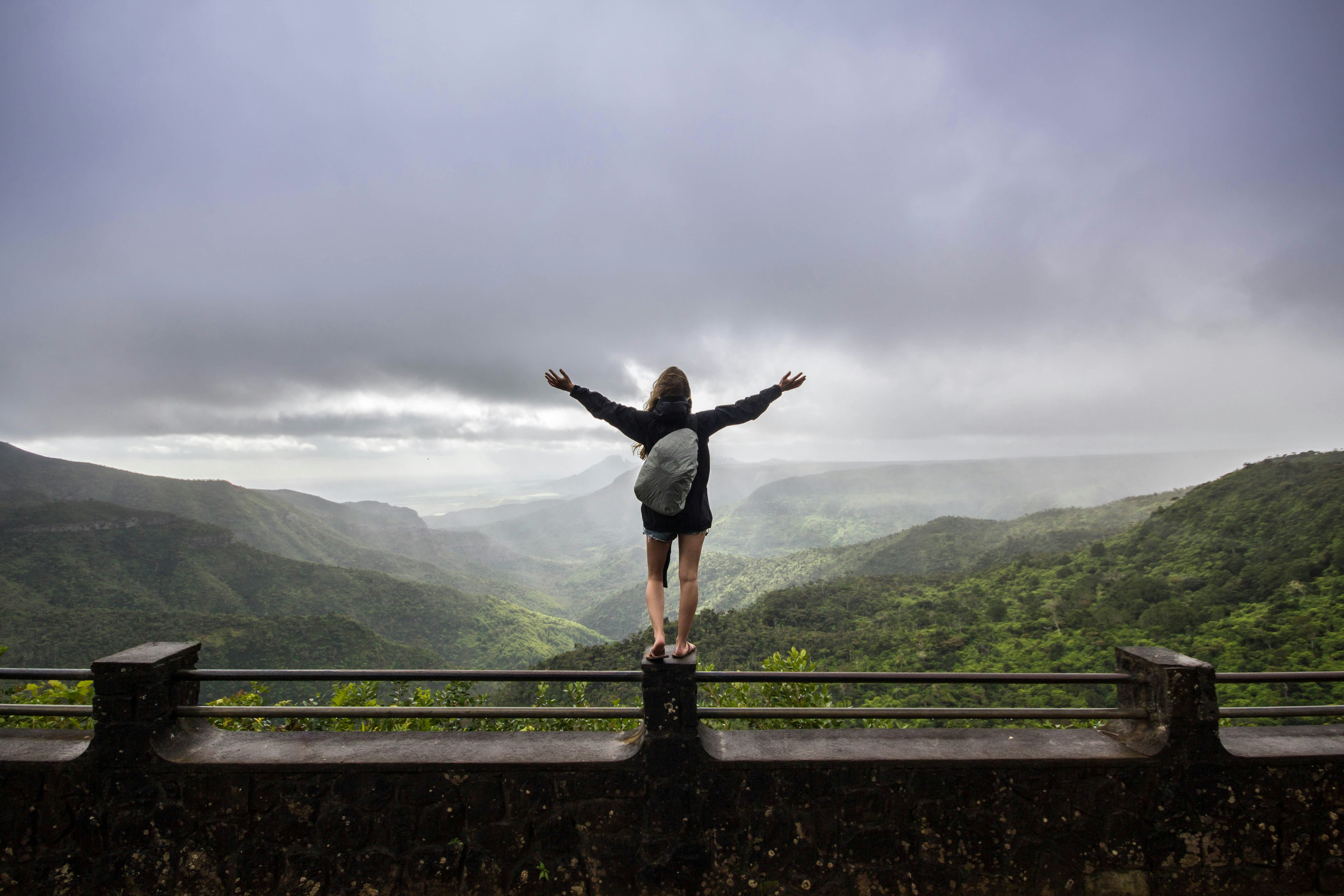Person standing on a ledge with arms outstretched, overlooking a lush green valley under a moody sky.