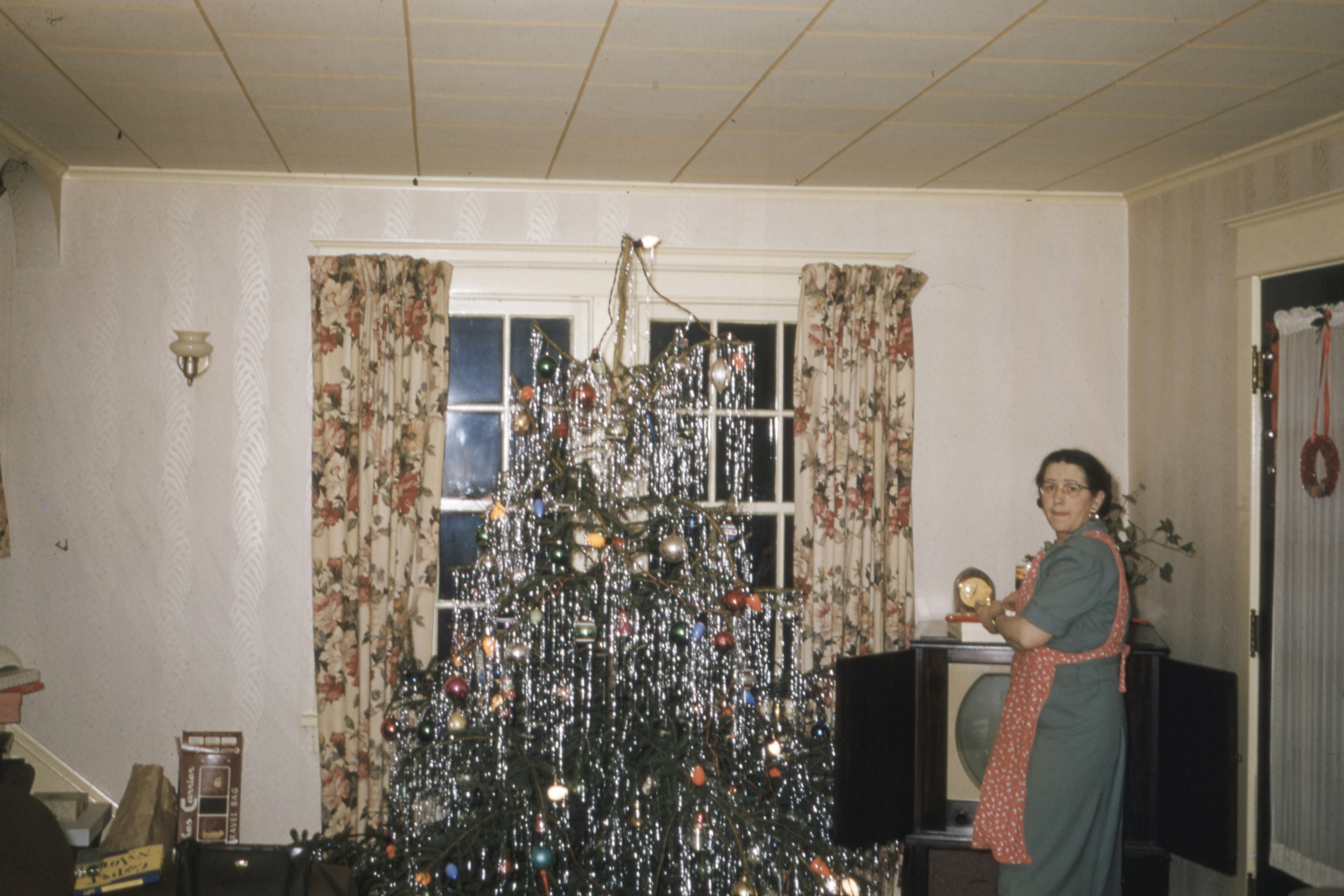 A candid photograph of a midcentury living room with a decorated Christmas tree in front of floral curtains, while a woman in a green dress stands to the right near a vintage console.