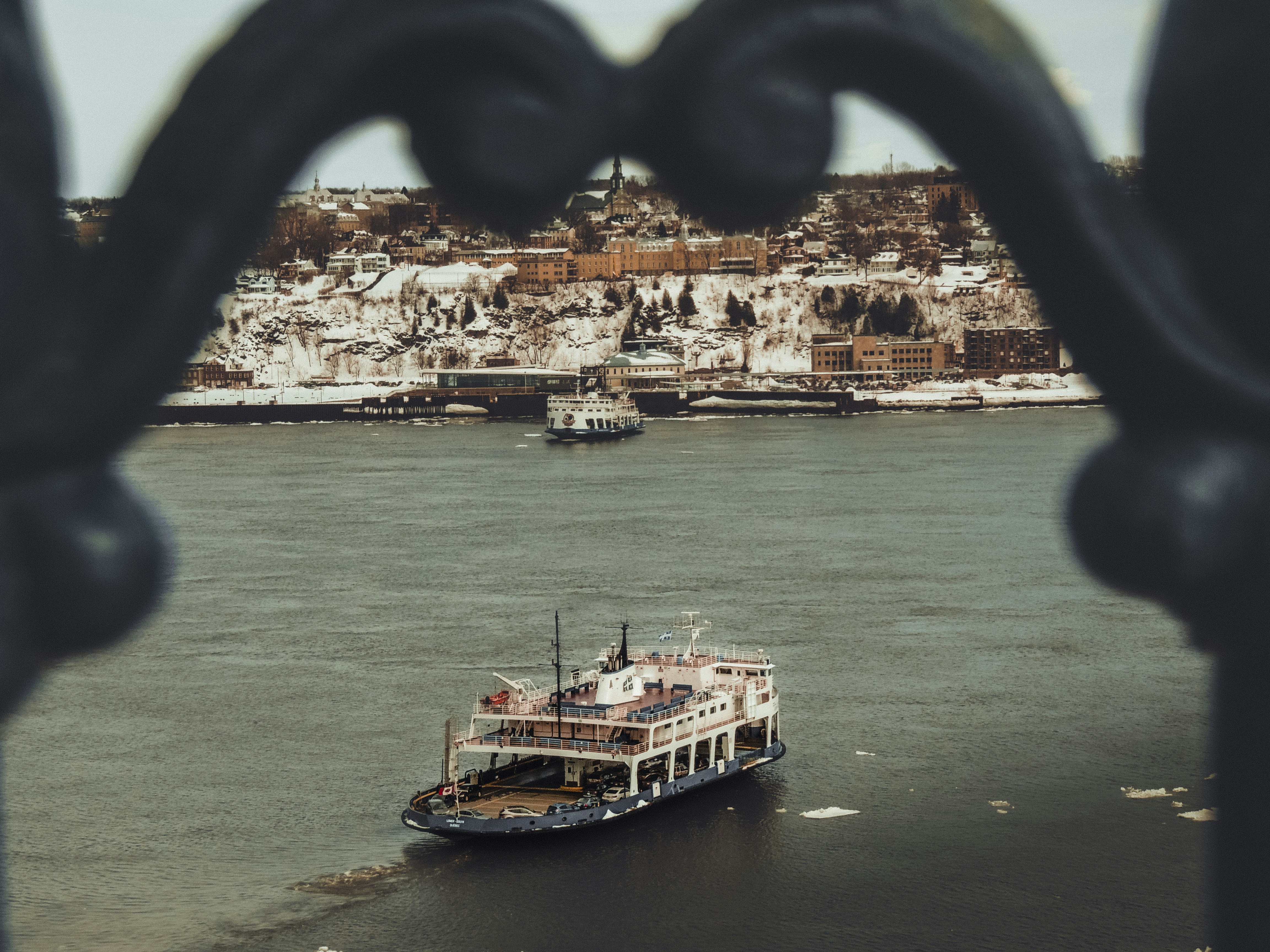 Ferry traversing icy waters, framed by ornate iron railing with snowy hillside in the background.