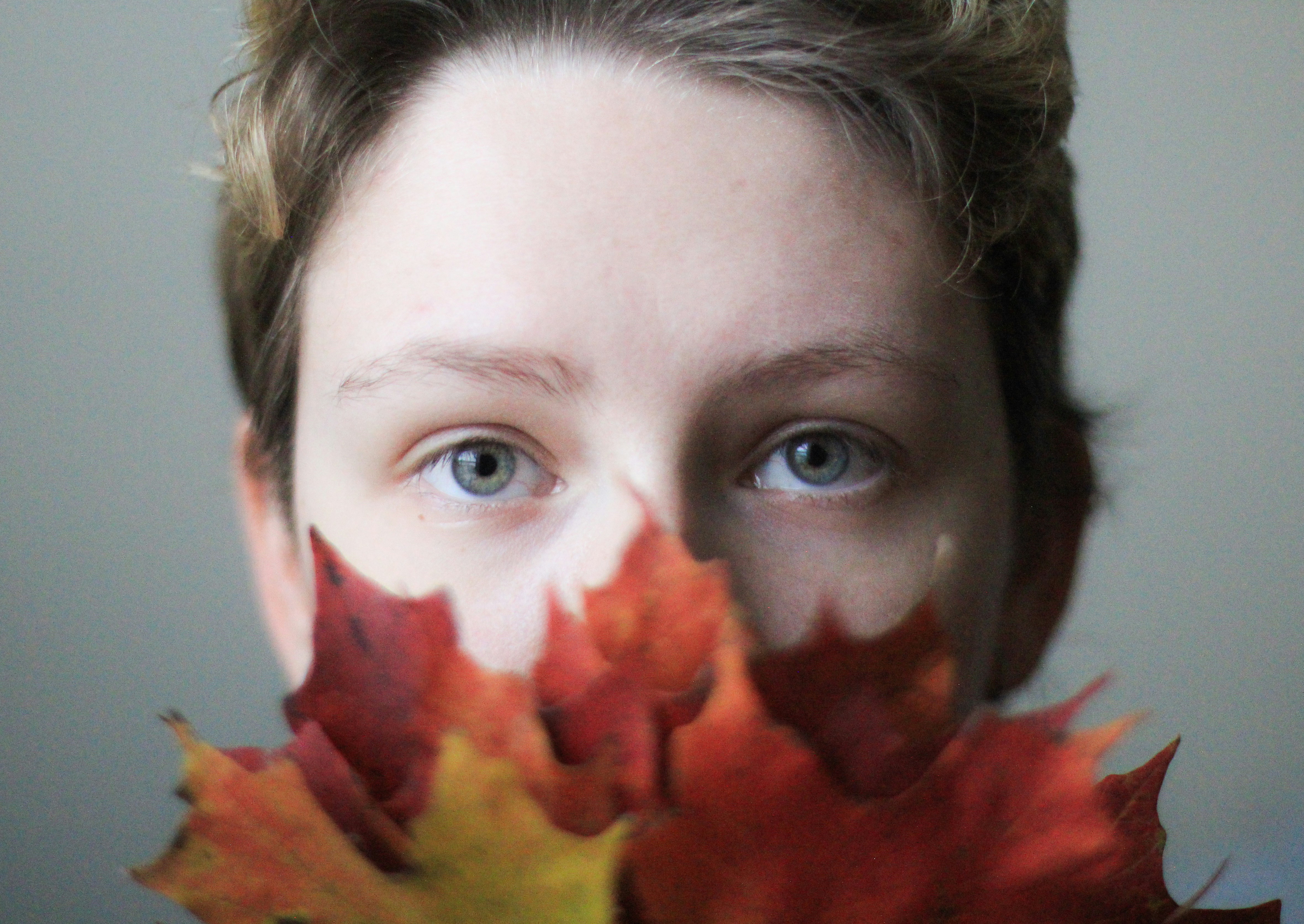 A person partially obscured by vibrant autumn leaves, with piercing blue eyes visible. The composition highlights the interplay between human expression and nature.