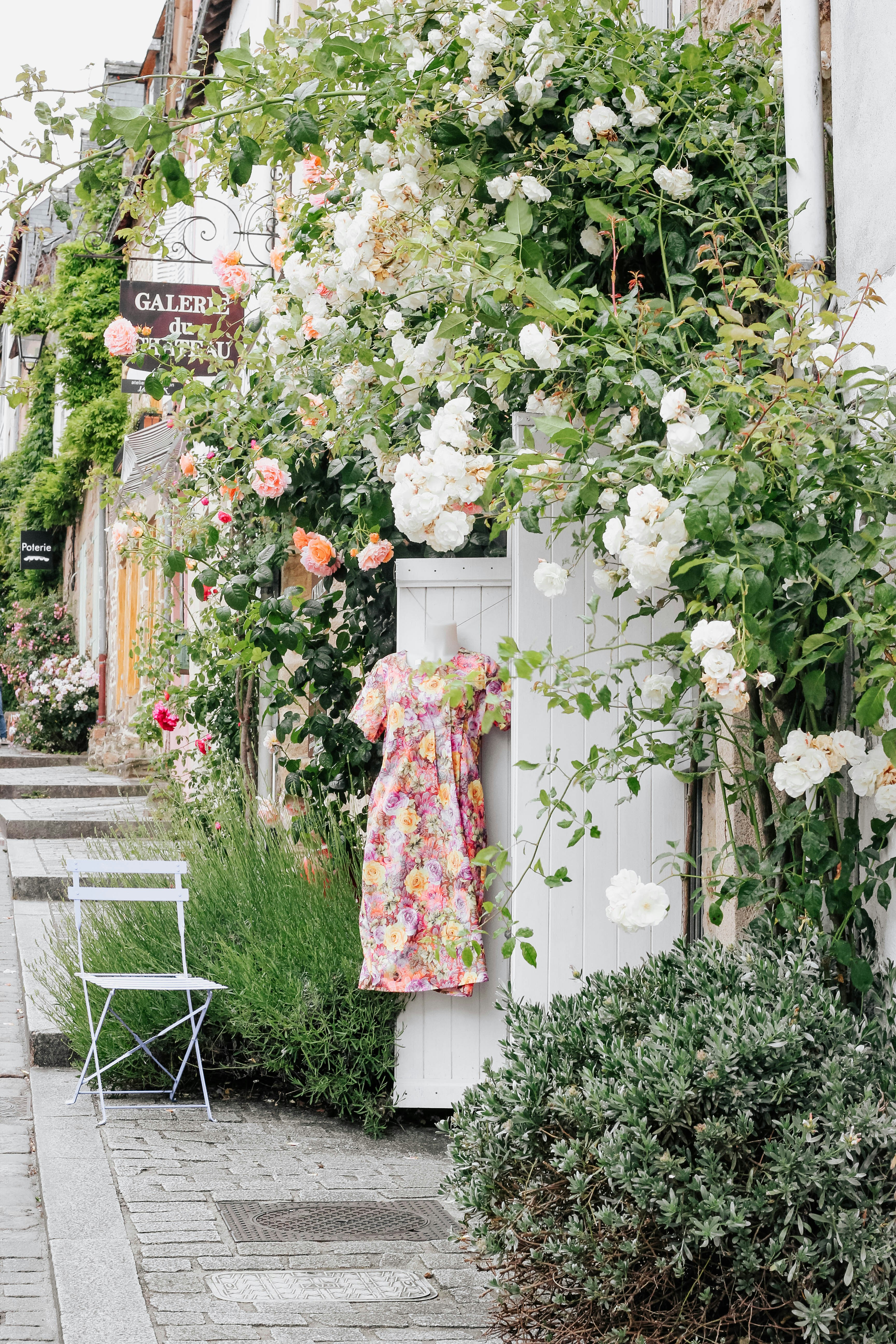 A vibrant floral dress hangs against a white wooden fence, surrounded by blooming roses along a quaint cobblestone street. A small chair adds to the charm of this picturesque scene.