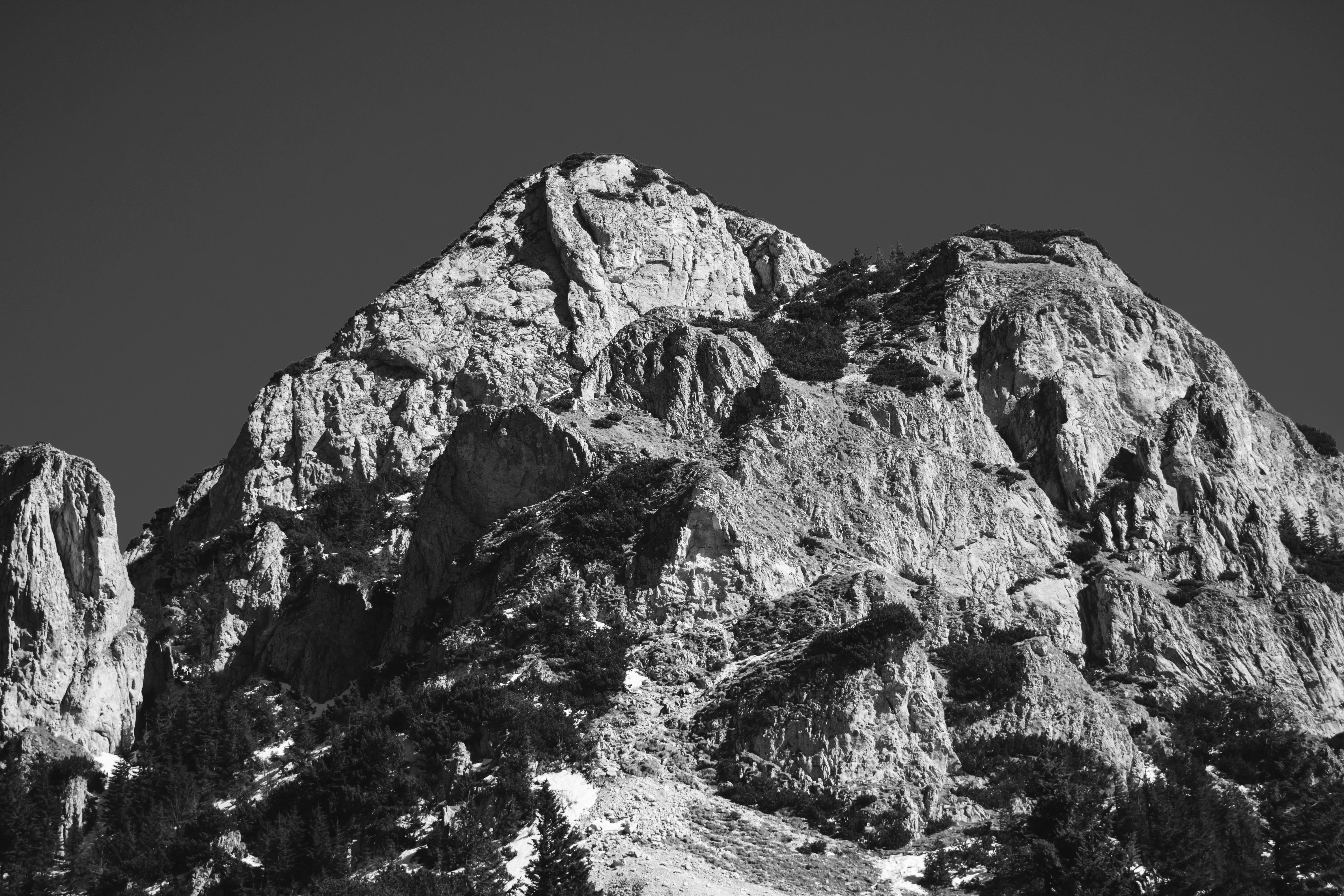 A rugged mountain peak rises dramatically against a clear sky, showcasing its textured rock face and surrounding terrain in striking black and white.