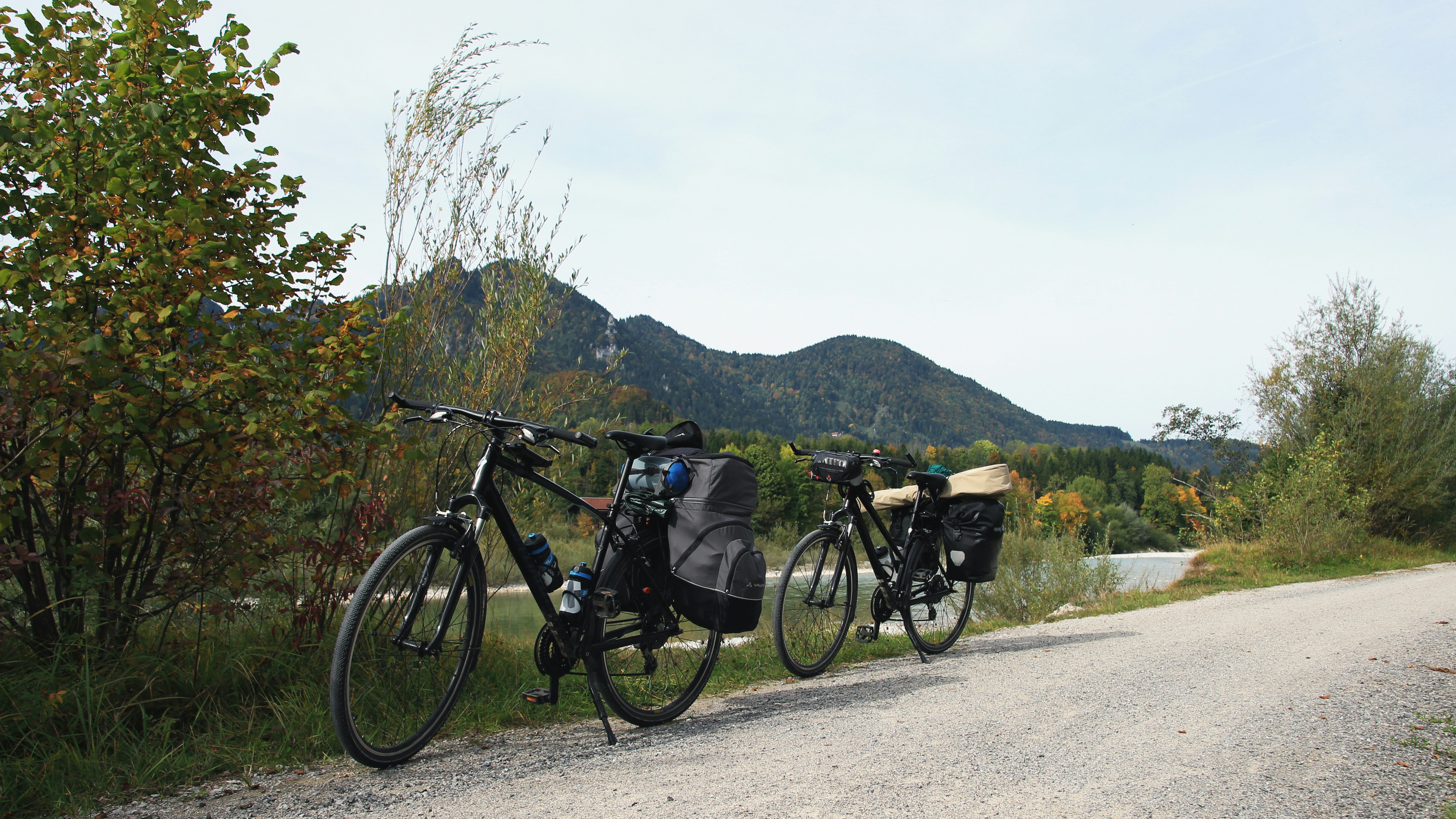 Two mountain bikes rest on a gravel path beside lush greenery and distant peaks during the day.