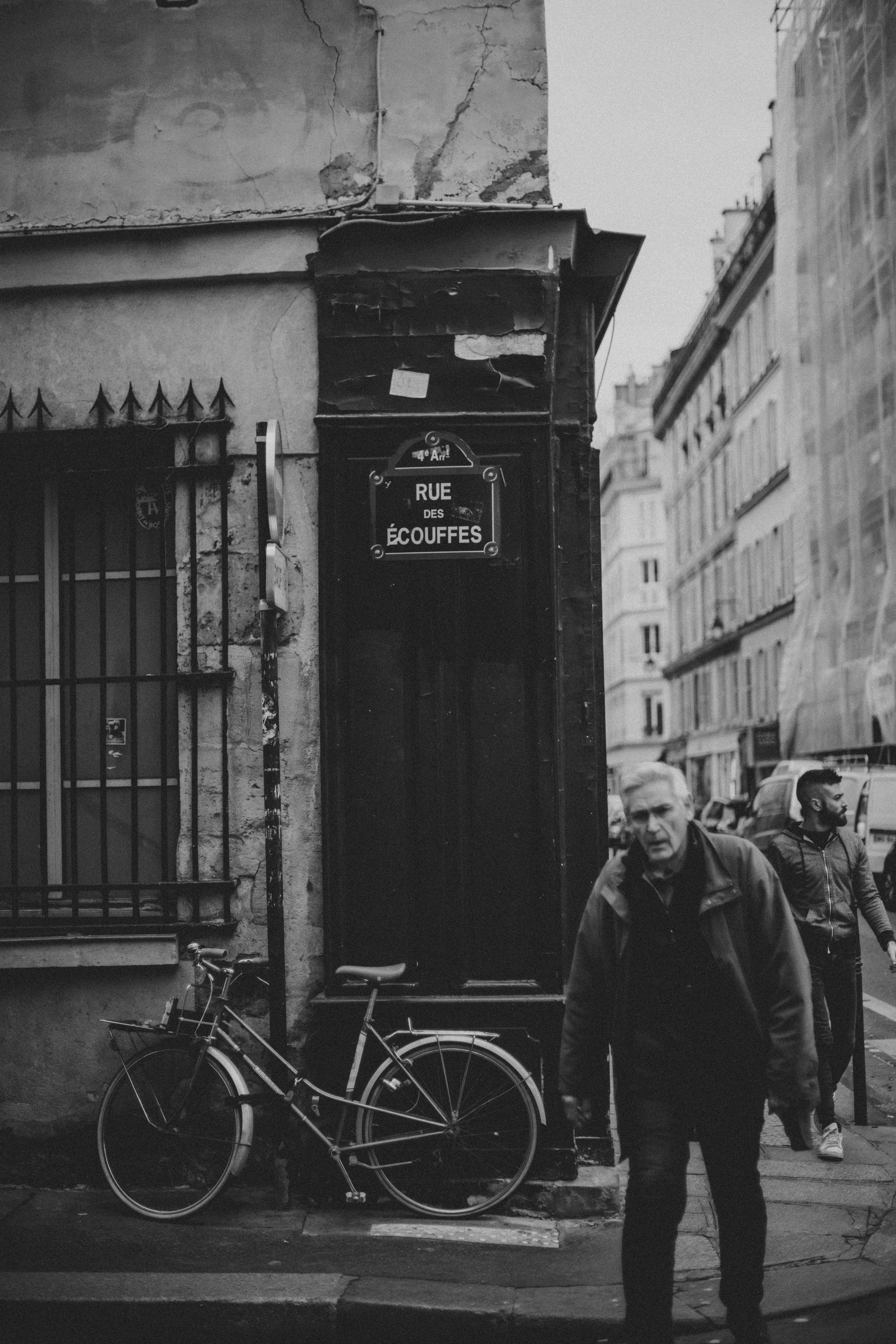 Monochrome Corner Moment on Rue des Écouffes: A Pedestrian, a Bicycle, and Weathered Facades