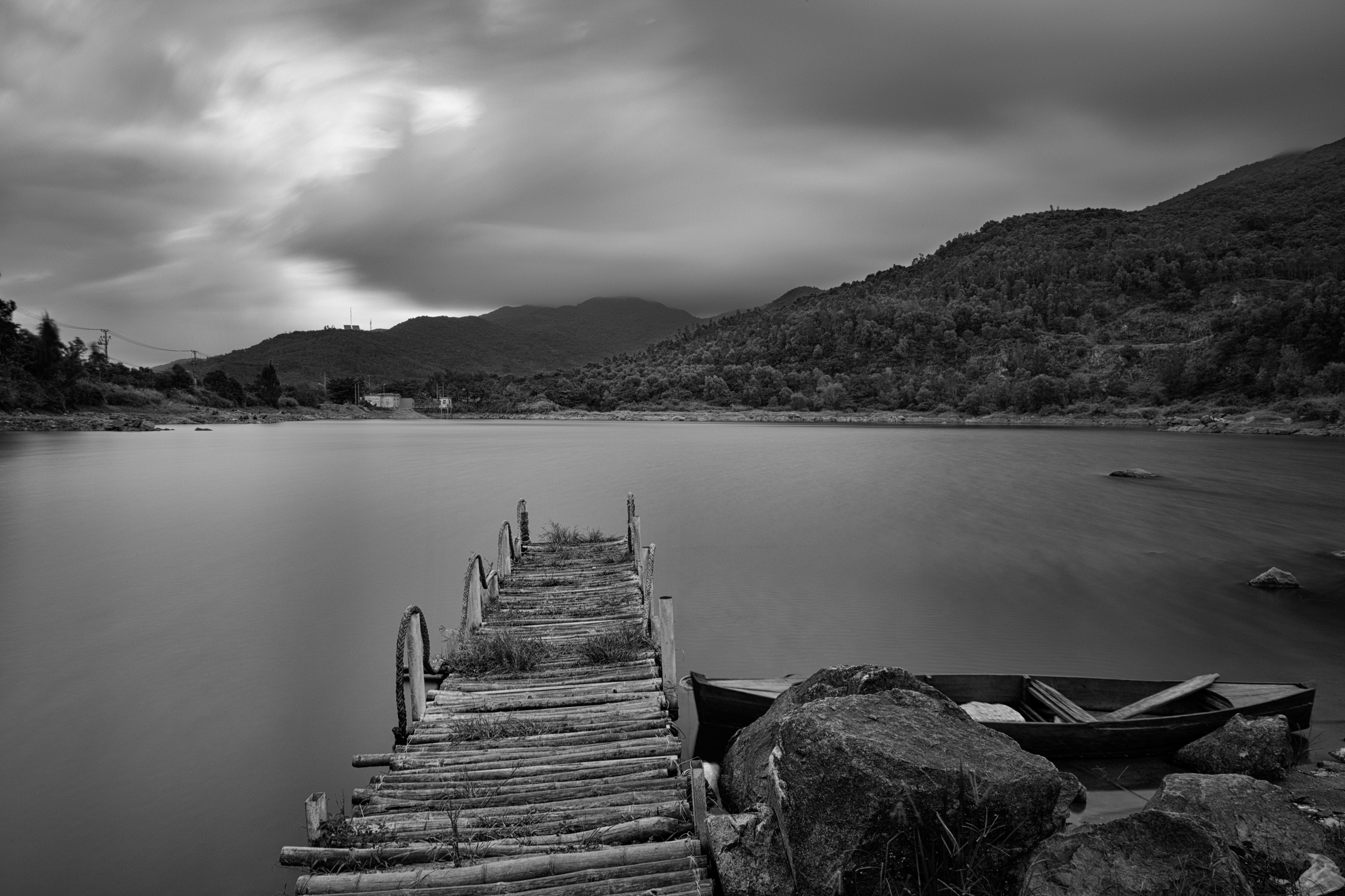 A dilapidated wooden dock extends into a tranquil lake under a cloudy sky, flanked by distant hills.