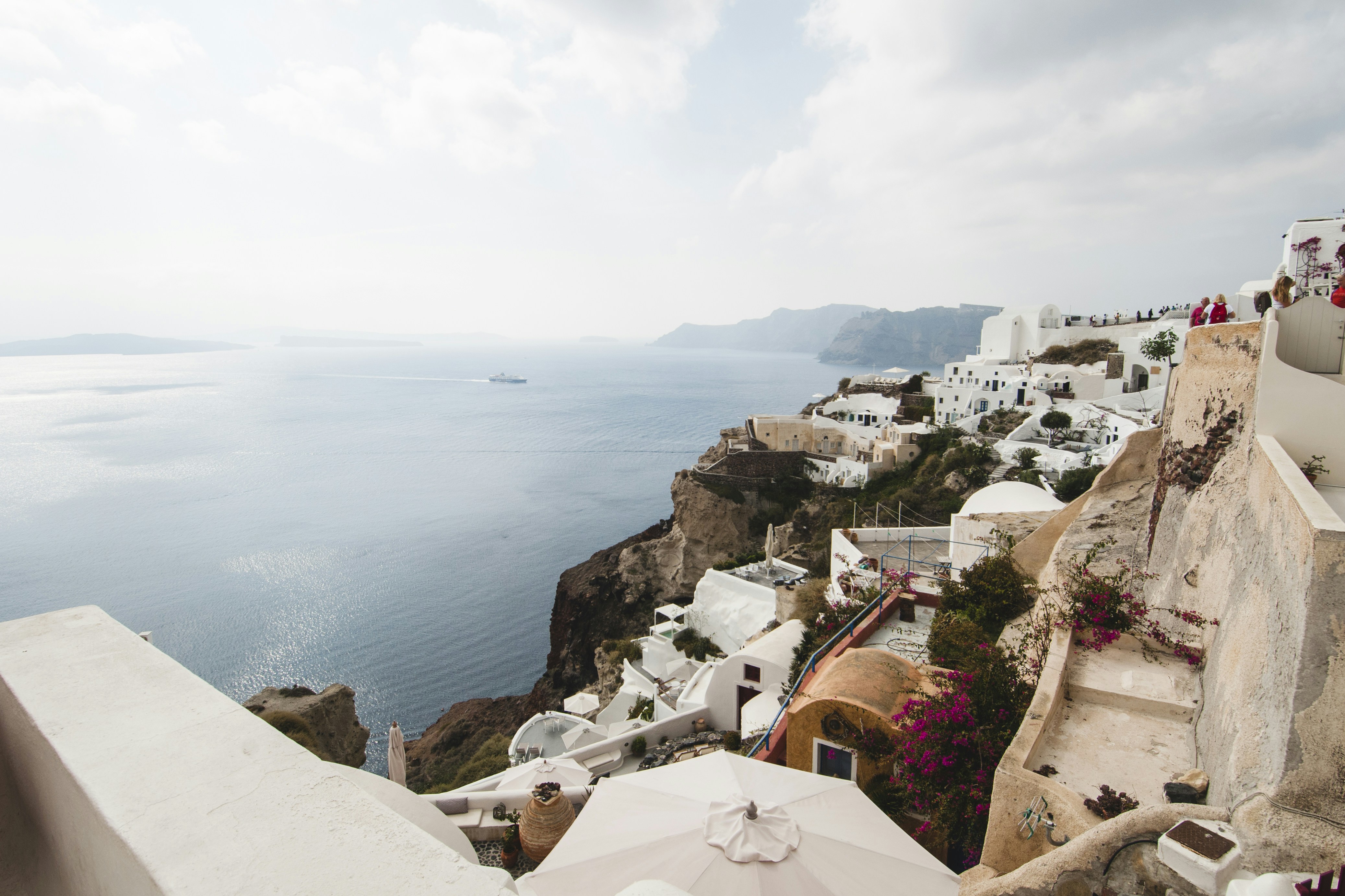 Whitewashed buildings perched on a cliffside overlooking the tranquil Aegean Sea under a hazy sky.