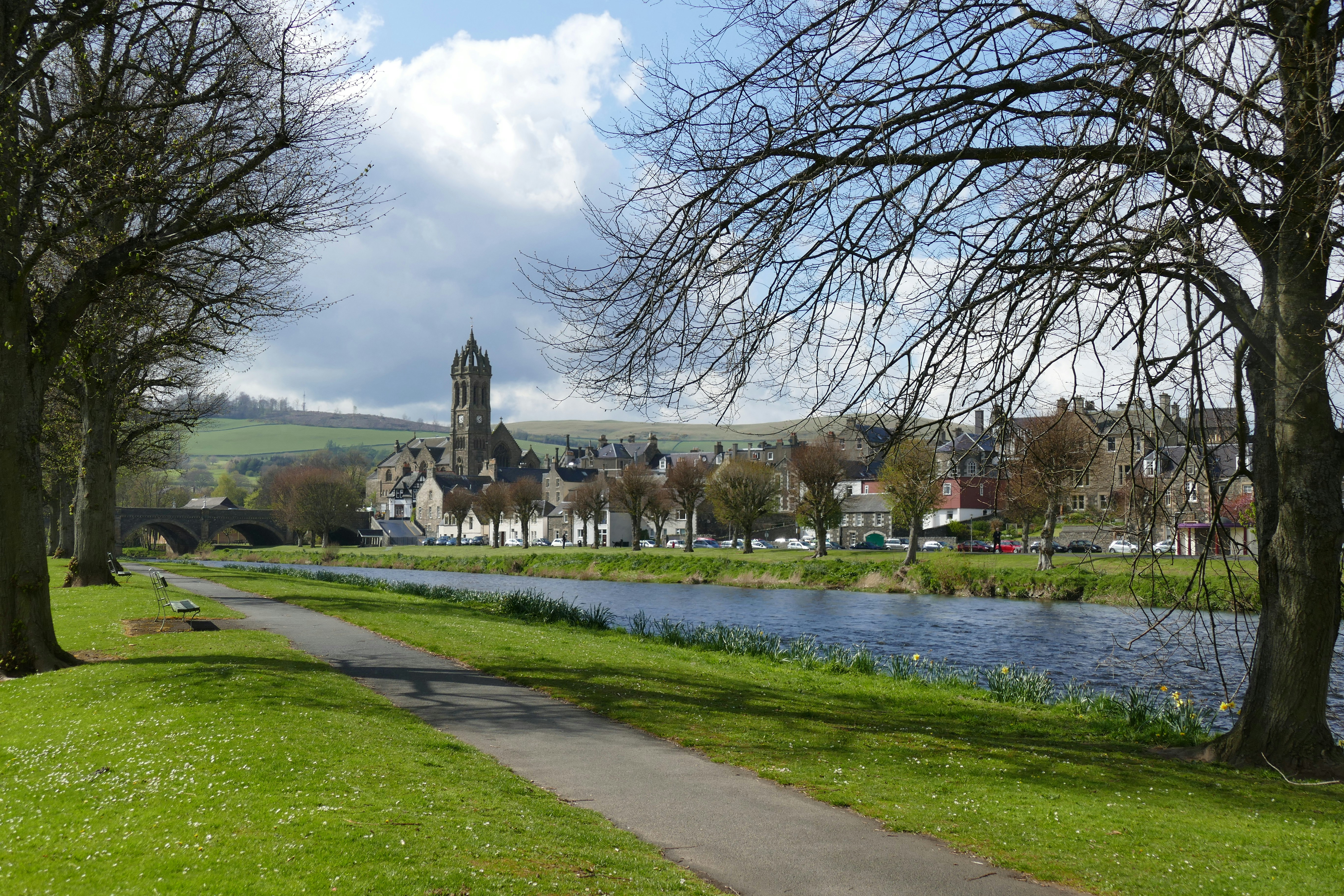 Pathway along a river in Peebles with a historic tower and distant hills under a partly cloudy sky.