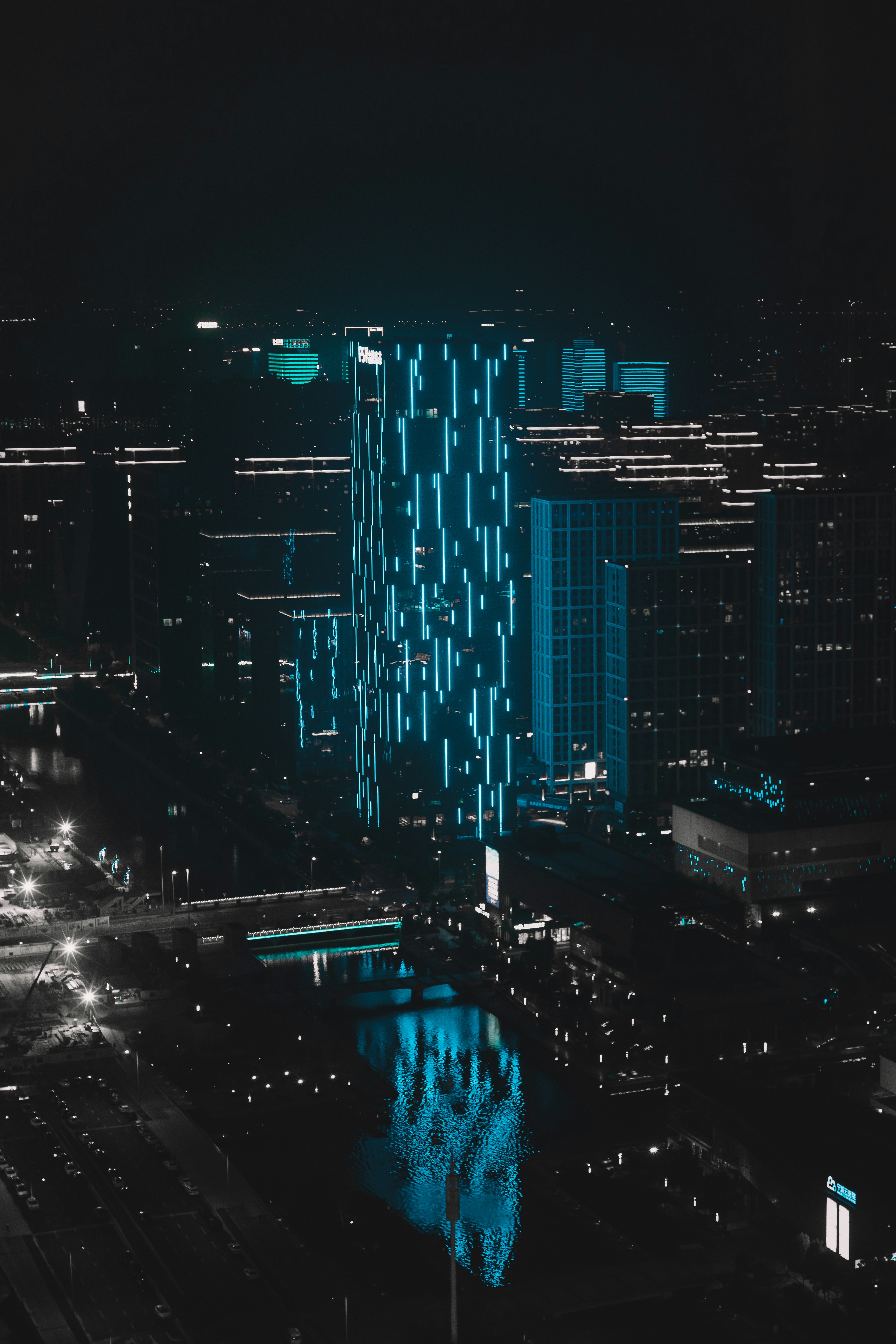 Illuminated skyscraper adorned with vertical blue lights, surrounded by a bustling cityscape at night, reflecting in the water below.