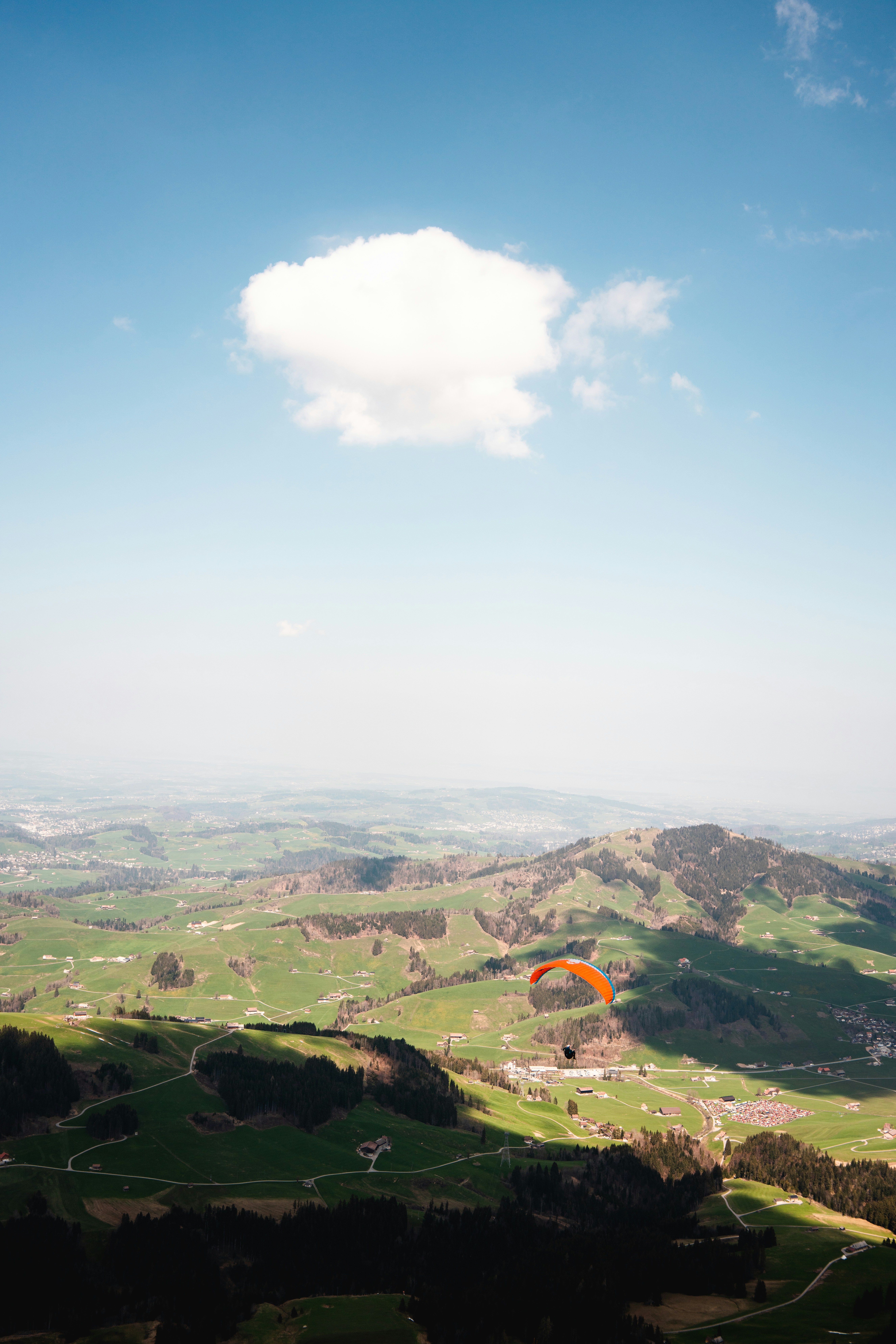 Paraglider glides over a patchwork of green hills under a pale blue sky with a single cloud above a distant horizon. The scene captures expansive terrain and a hint of human activity within the natural landscape.