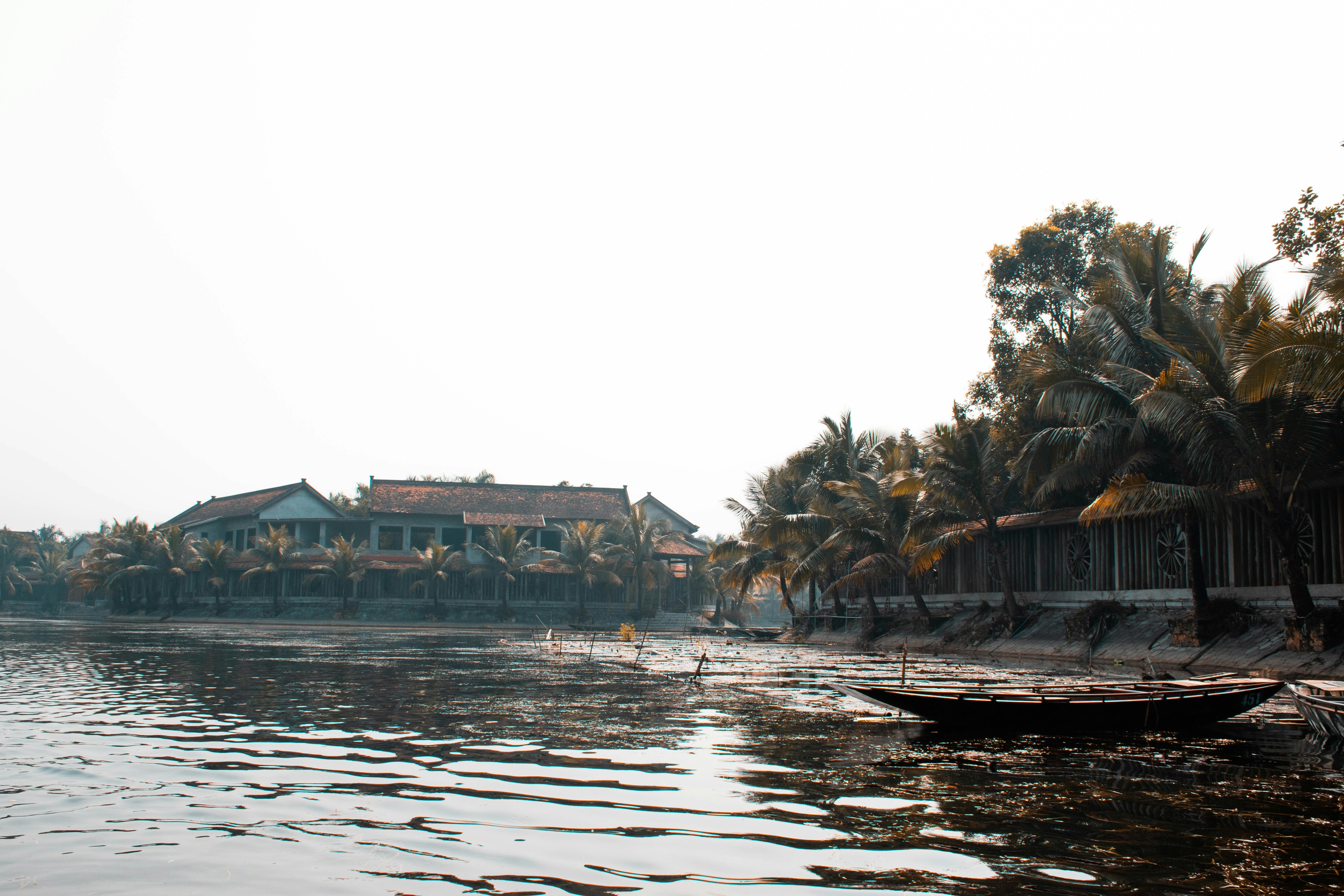 Wooden boats rest on a tranquil shore lined with coconut trees under a hazy sky.
