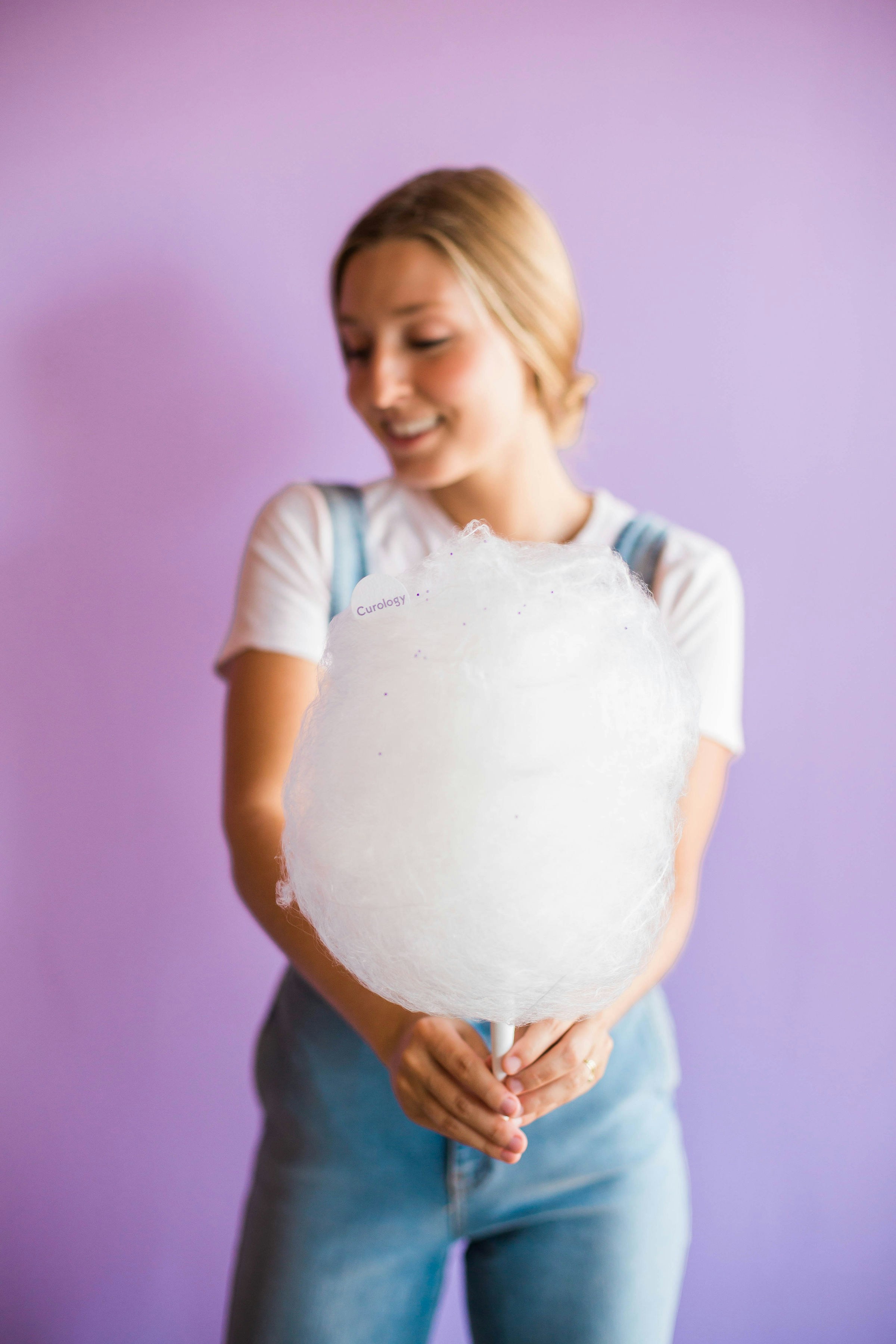 A young woman joyfully holds a large cotton candy against a soft purple backdrop, embodying the spirit of childhood nostalgia.