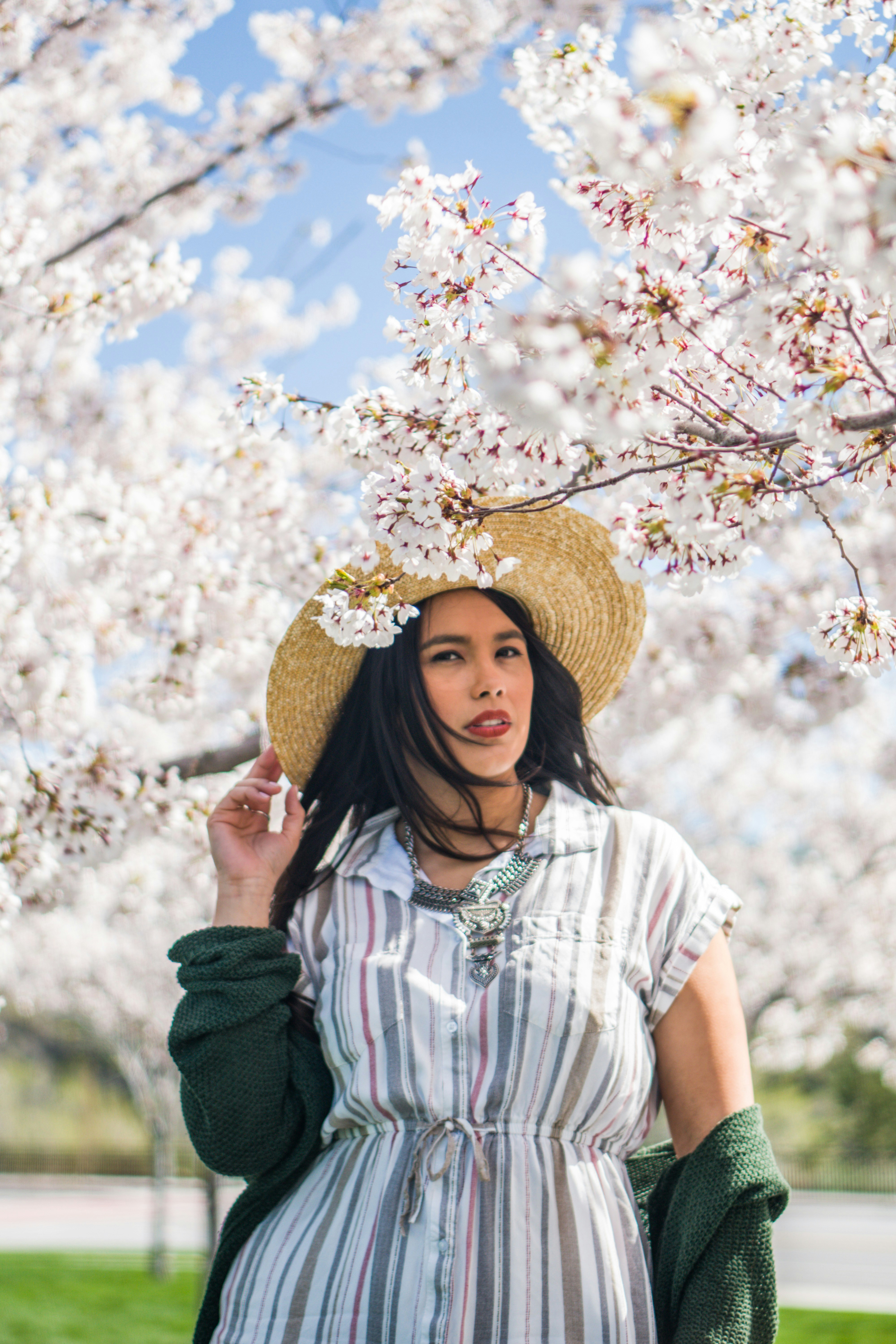 A woman in a straw hat poses among blooming cherry blossoms, radiating joy and warmth. The vibrant colors and floral backdrop enhance the scene's lively atmosphere.