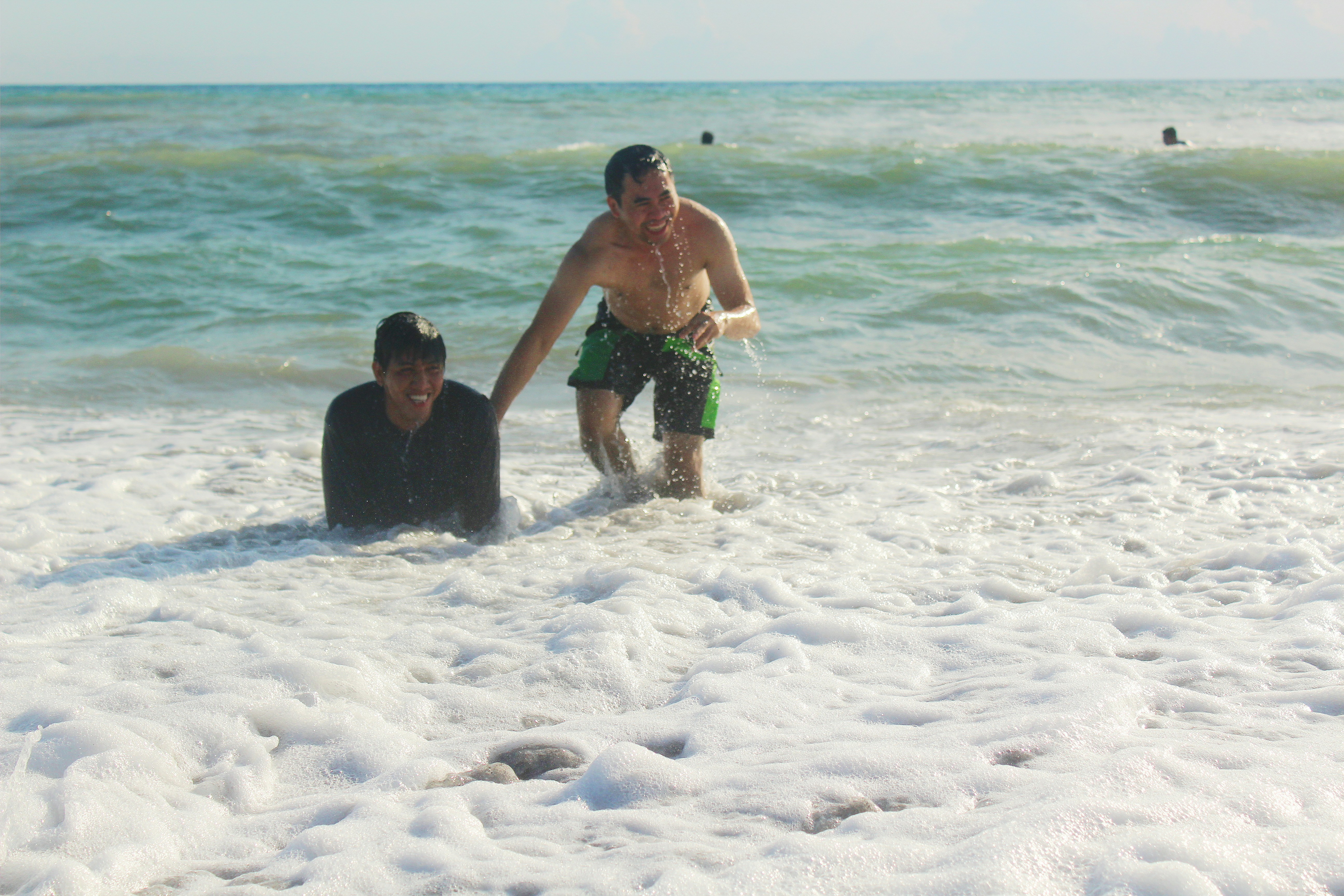 Two friends playfully interacting in the surf, with foamy waves crashing around them. The scene captures a lively beach atmosphere.
