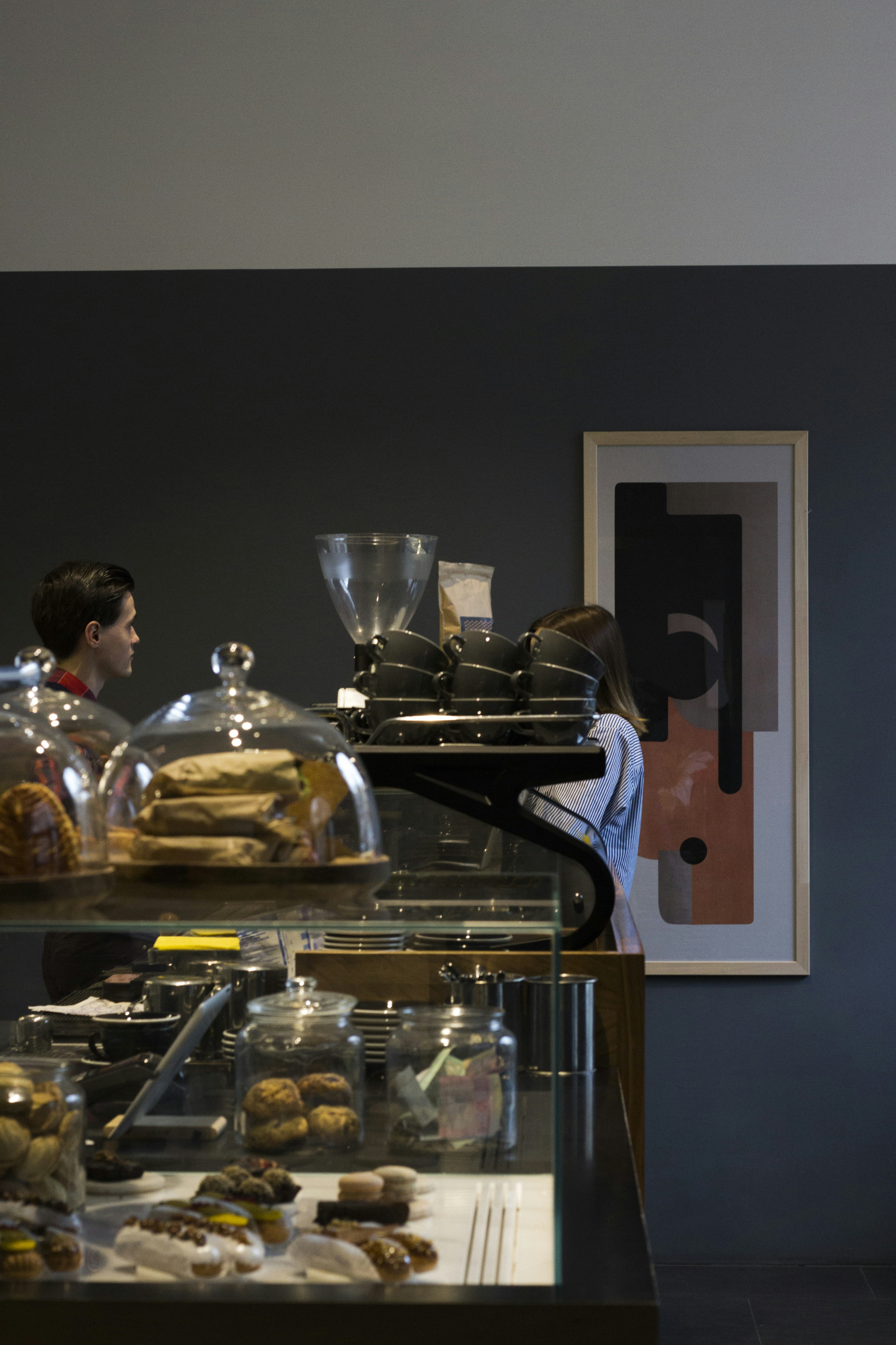A sleek café interior featuring a barista and a customer, with a striking abstract artwork on the wall and an array of pastries displayed in glass cases.