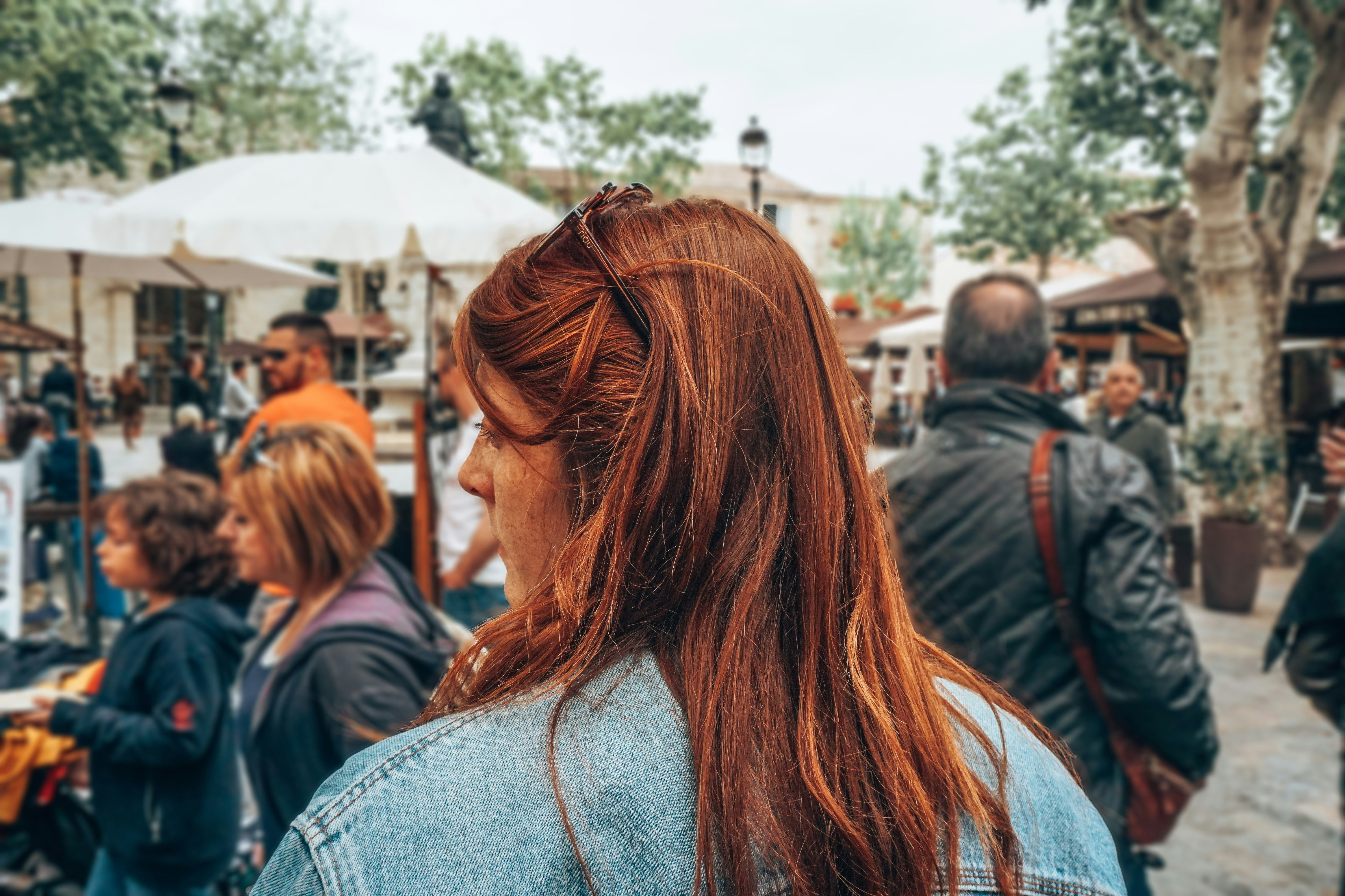 Woman with long red hair in a denim jacket stands amidst a bustling market scene filled with people and outdoor cafes.