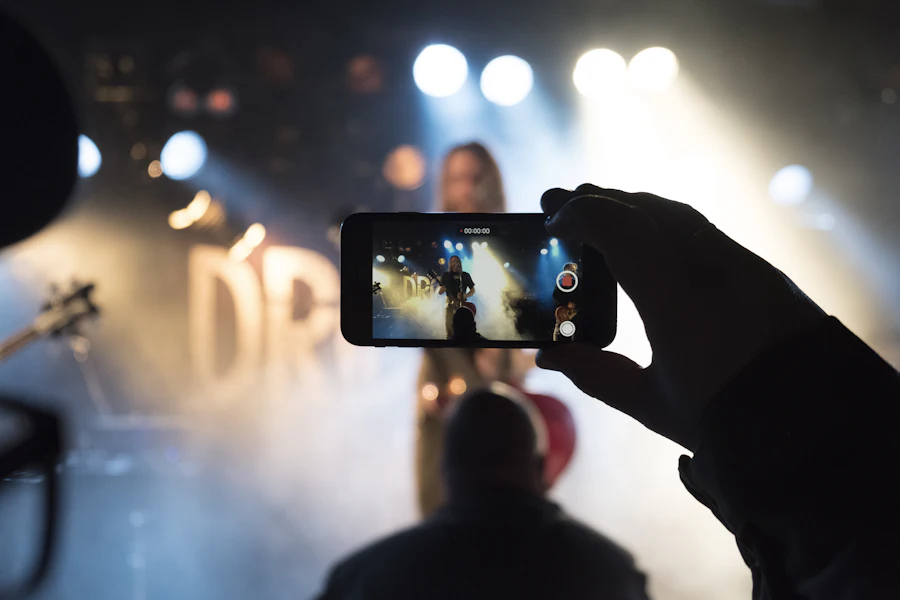 A hand holds up a smartphone recording a singer on a smoky stage, with bright spotlights and blurred concertgoers in the foreground. The phone screen mirrors the performance as the crowd watches.