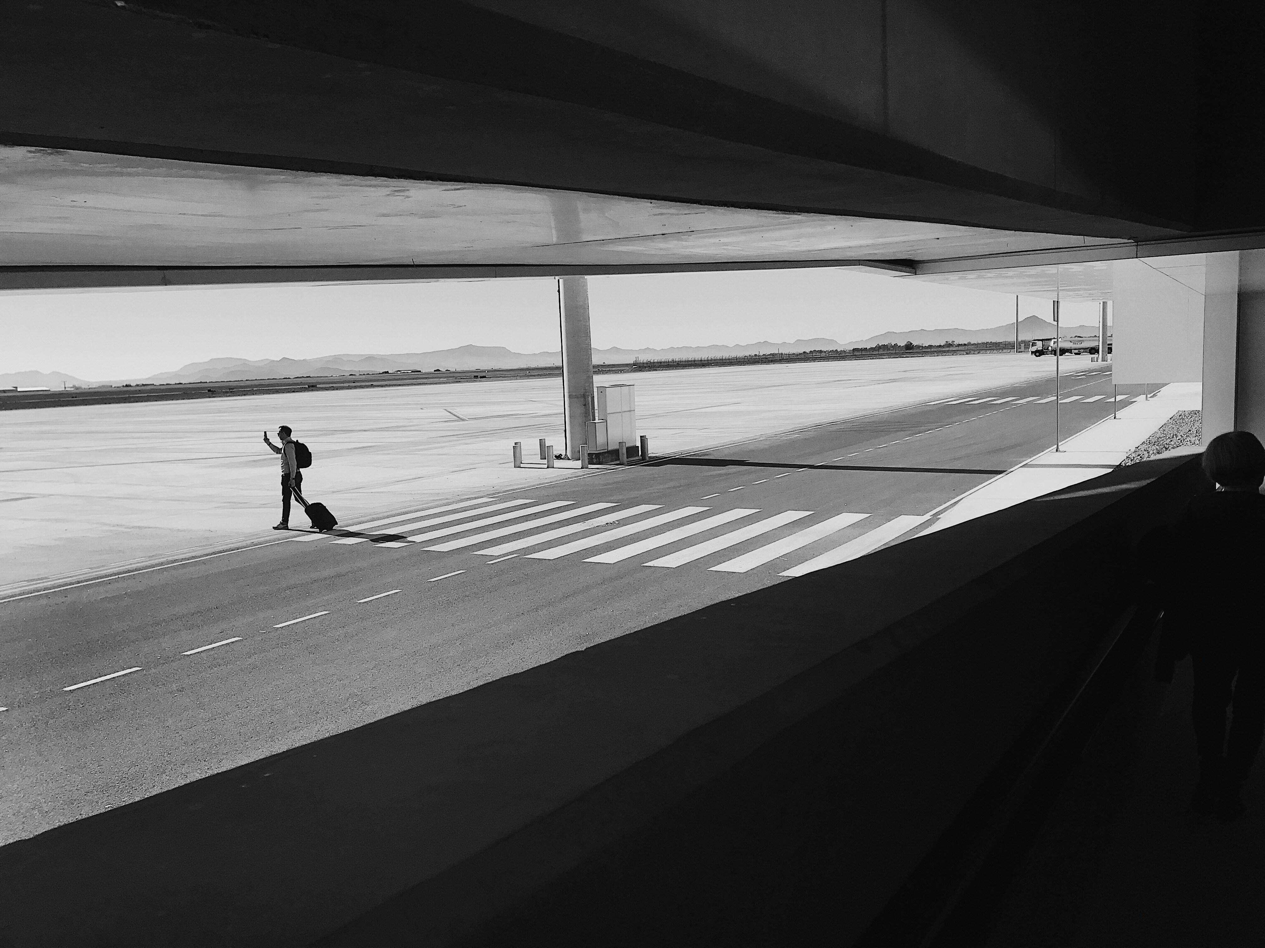 Silhouetted traveler pulling a suitcase across a sunlit airport tarmac framed by architectural shadows.