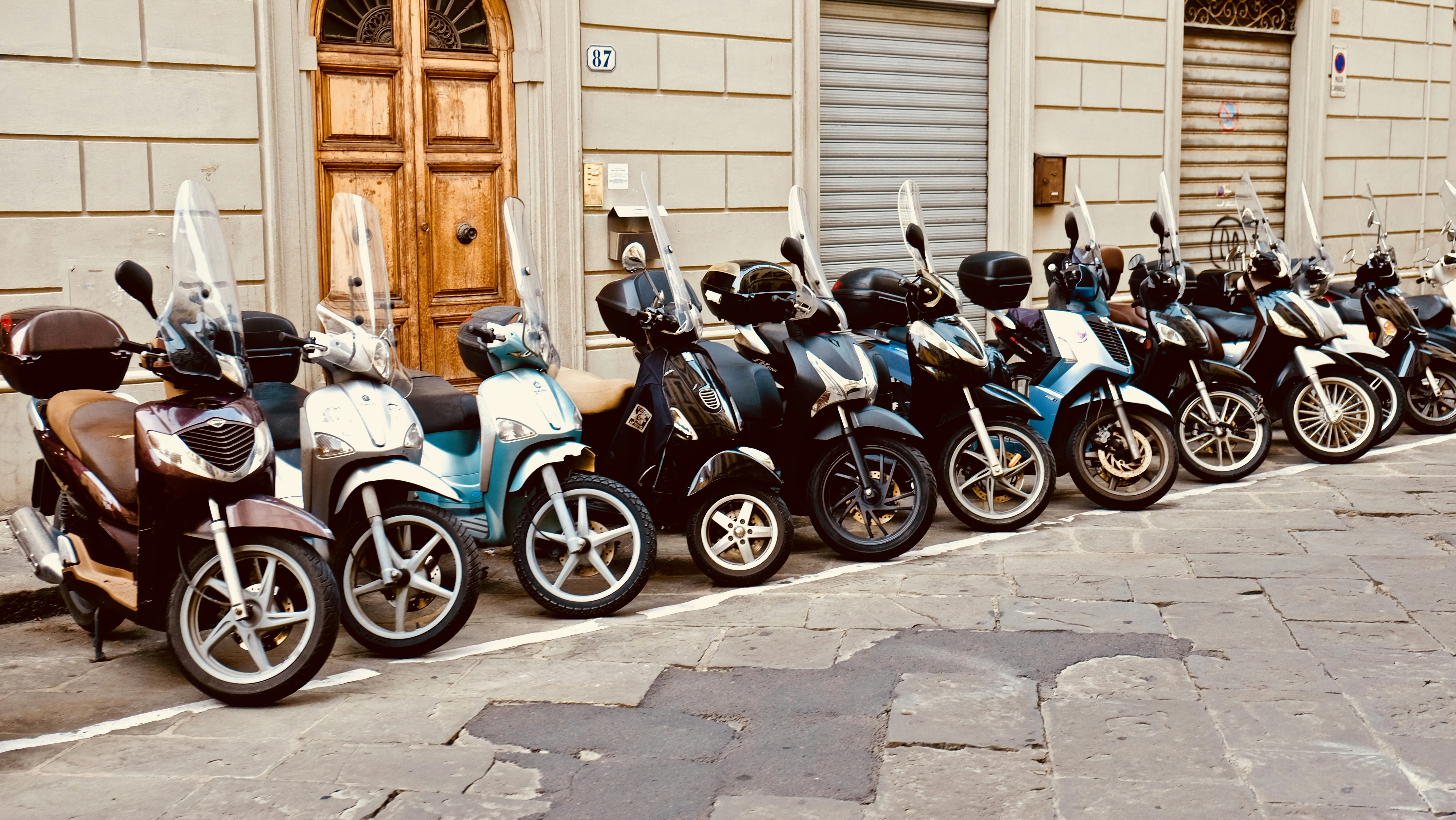 A row of scooters parked neatly against a textured wall, showcasing a variety of colors and styles. The wooden door in the background adds a rustic charm.