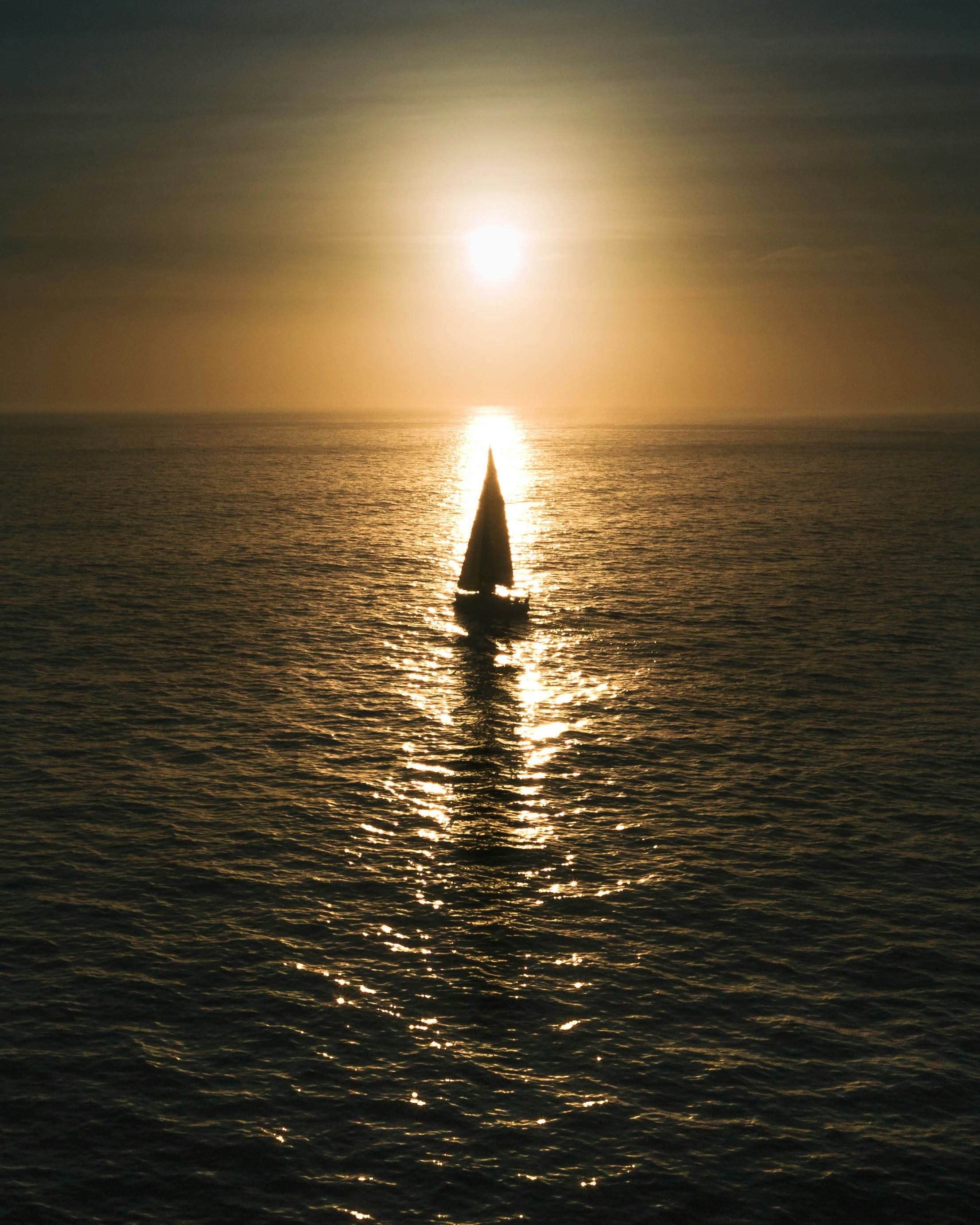 Silhouette of a sailboat gliding on tranquil waters as the sun sets, casting a golden reflection.