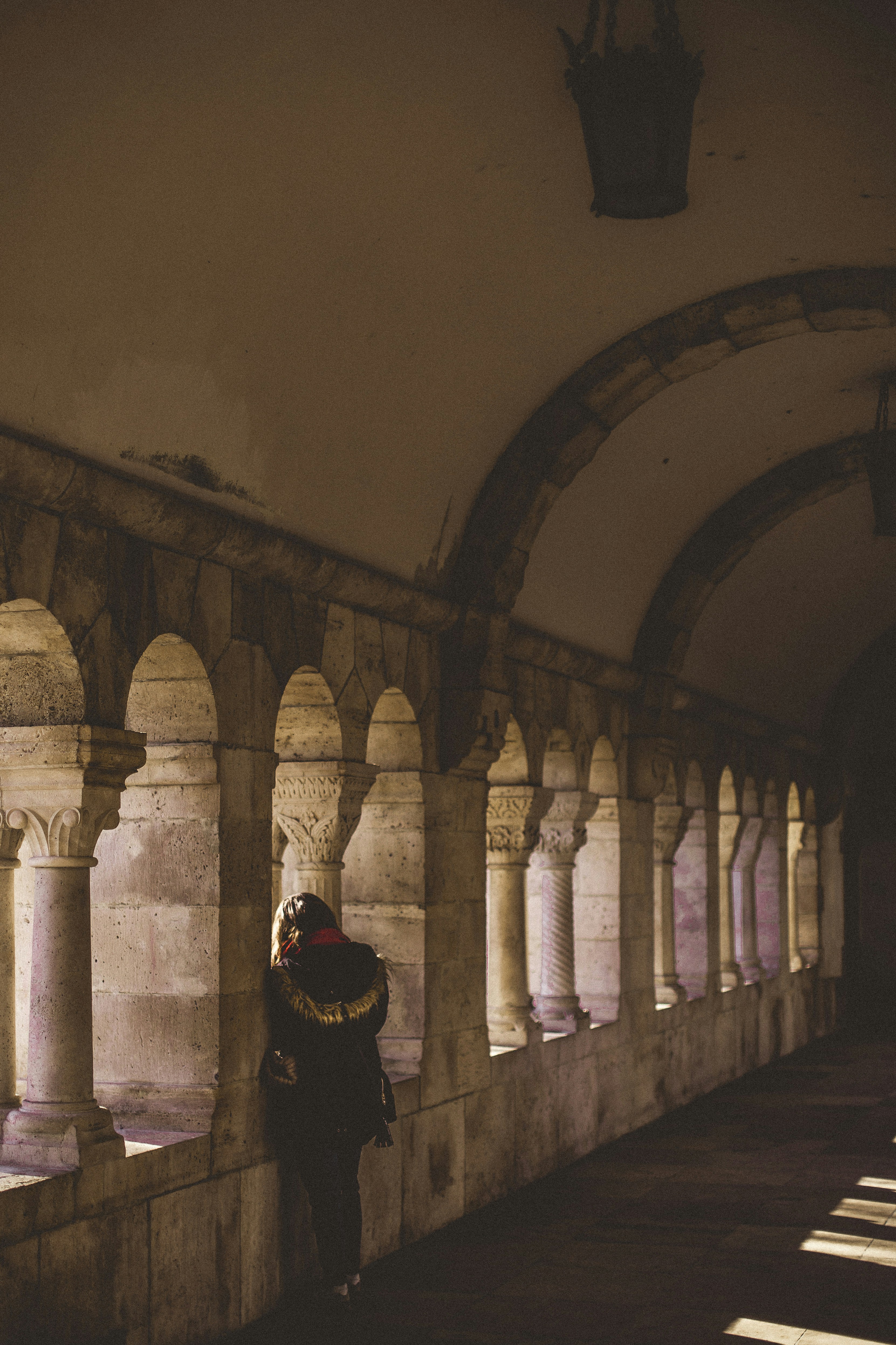 A solitary figure leans against a stone wall adorned with arches, illuminated by soft light filtering through the columns. The scene evokes a sense of contemplation and tranquility.