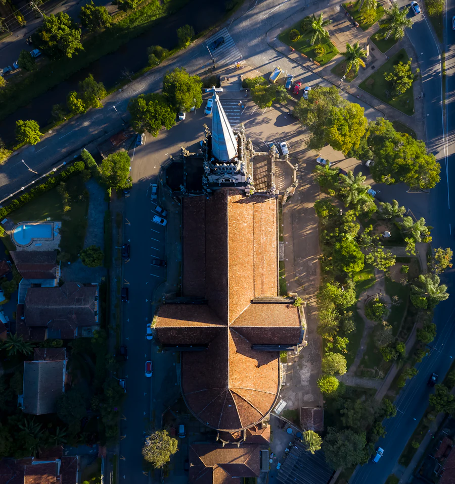 Top-down aerial view of a large church with terracotta roofs, a central spire, and surrounding tree-lined roads and paths. Long shadows stretch across the grounds in warm late-day light.