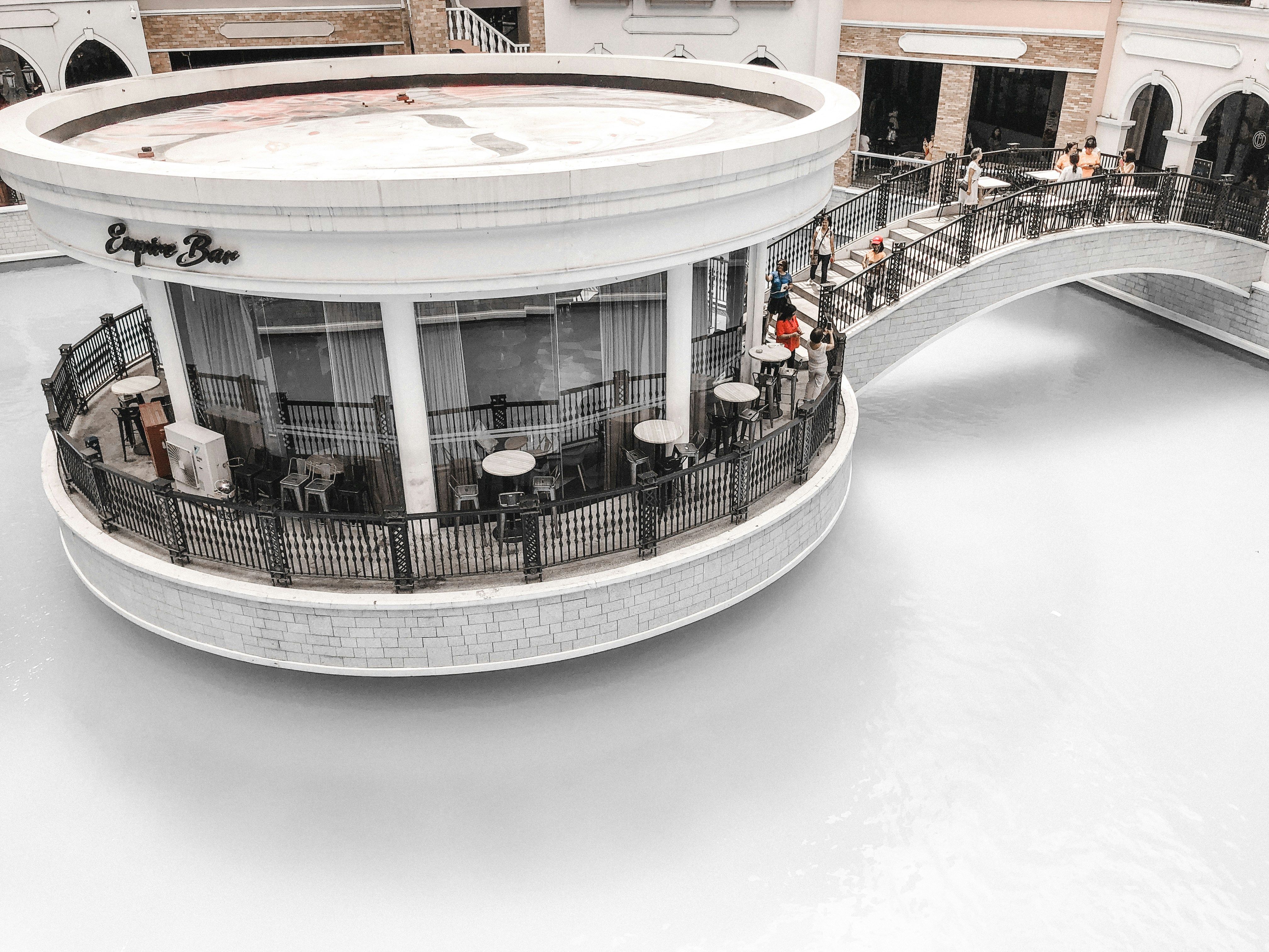 Circular café structure surrounded by tranquil waters, featuring outdoor seating and patrons enjoying their time. A bridge connects the café to the surrounding area.