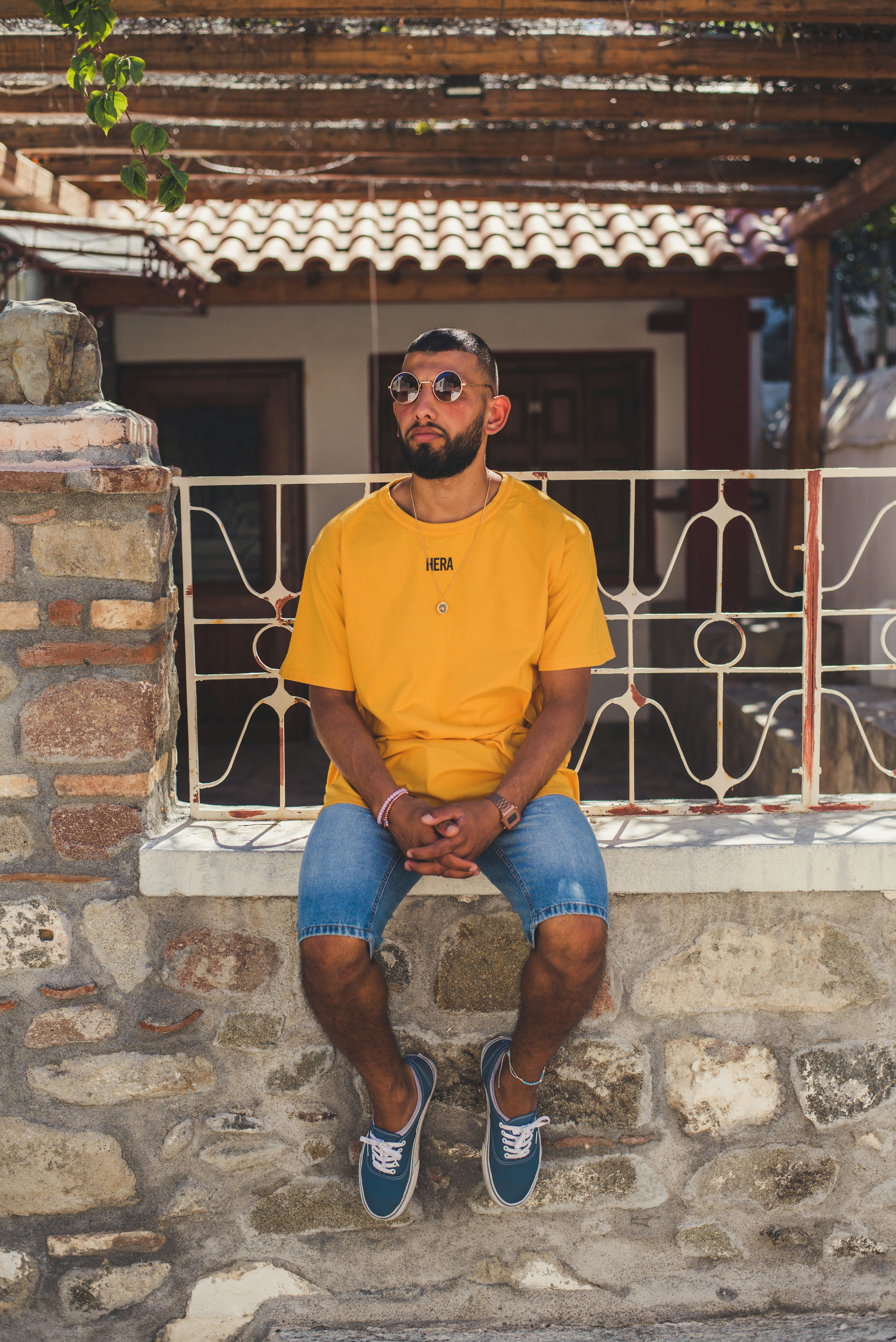 A man in a vibrant yellow shirt sits casually on a stone wall, framed by rustic architecture and shaded by a wooden pergola.