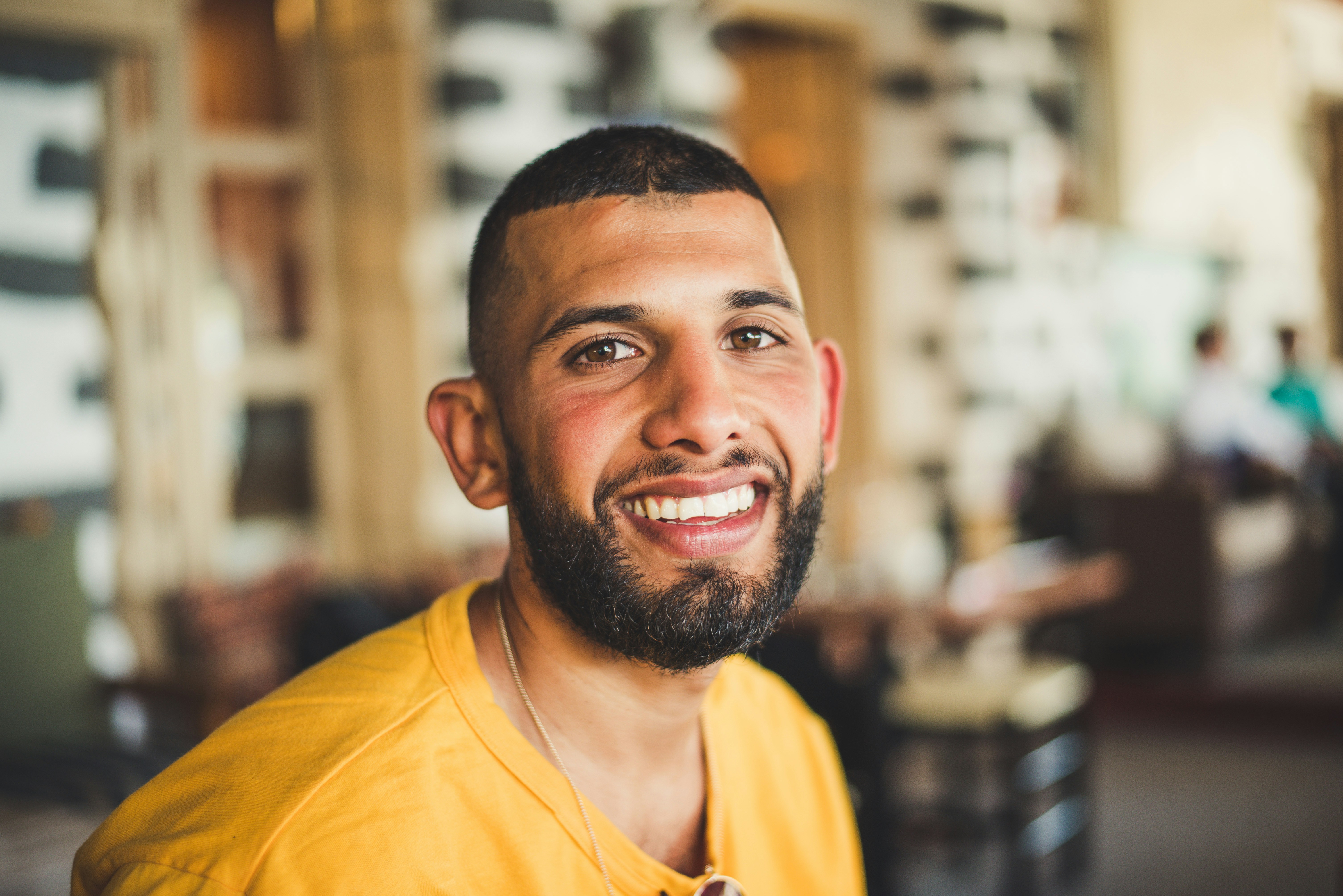 Smiling man in a yellow shirt, captured in a lively interior setting with soft lighting and blurred background elements. 
