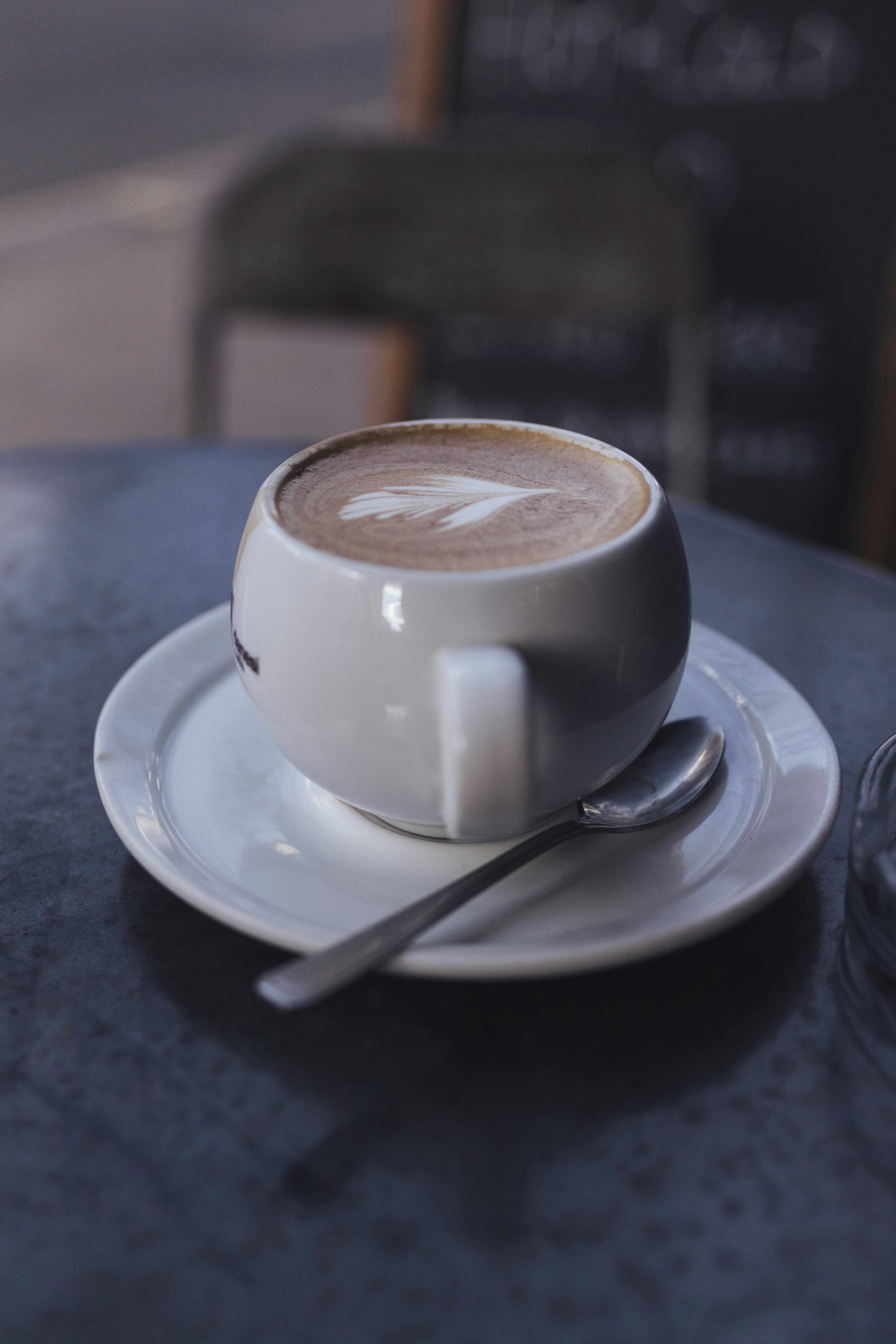 A carefully crafted cup of coffee sits on a saucer with a spoon, captured on a rustic table. The blurred background hints at a cozy café atmosphere.