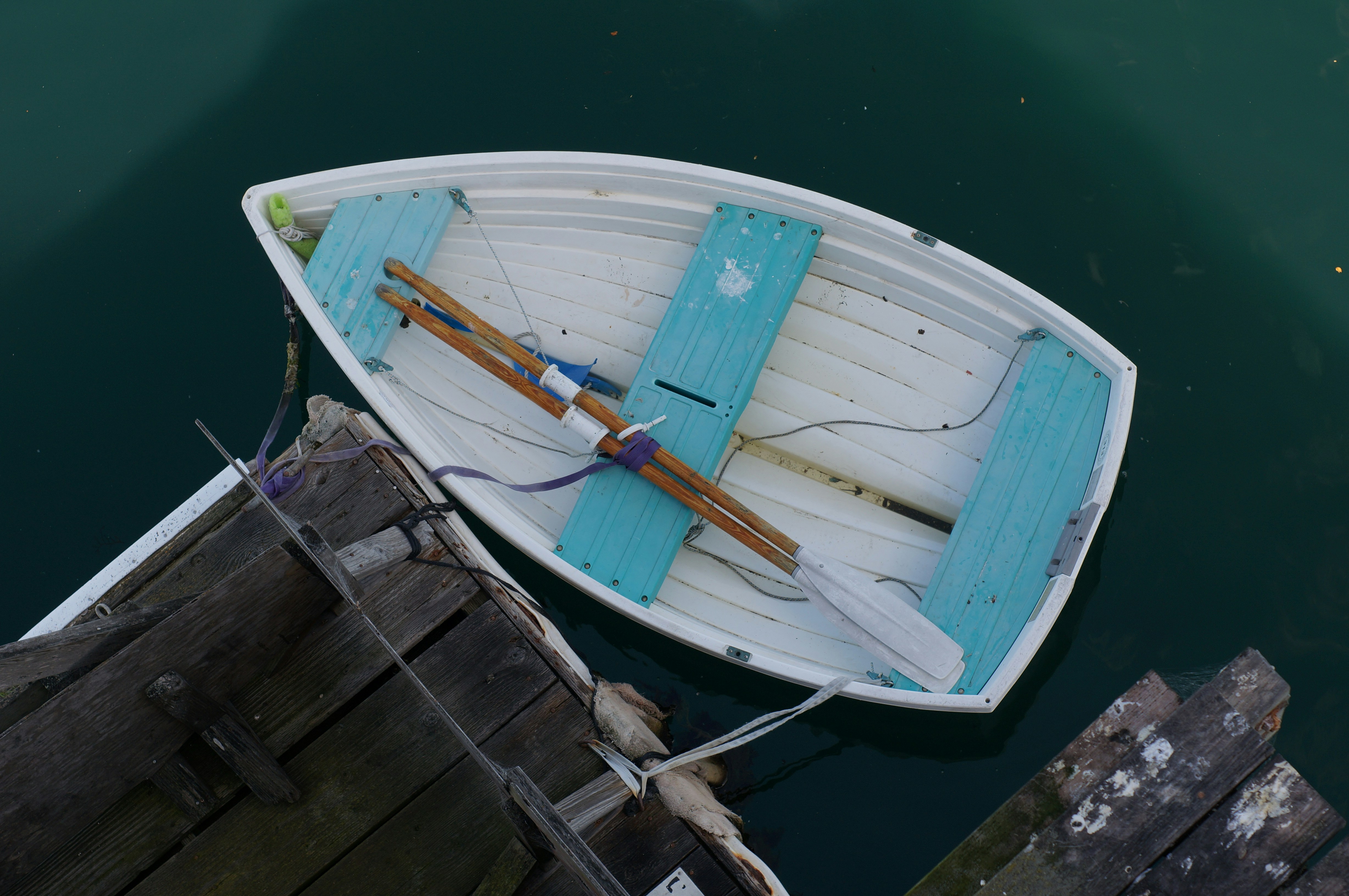 Top-down view of a white rowboat with turquoise seats and oars inside, tied to a weathered wooden dock, floating in calm green water.