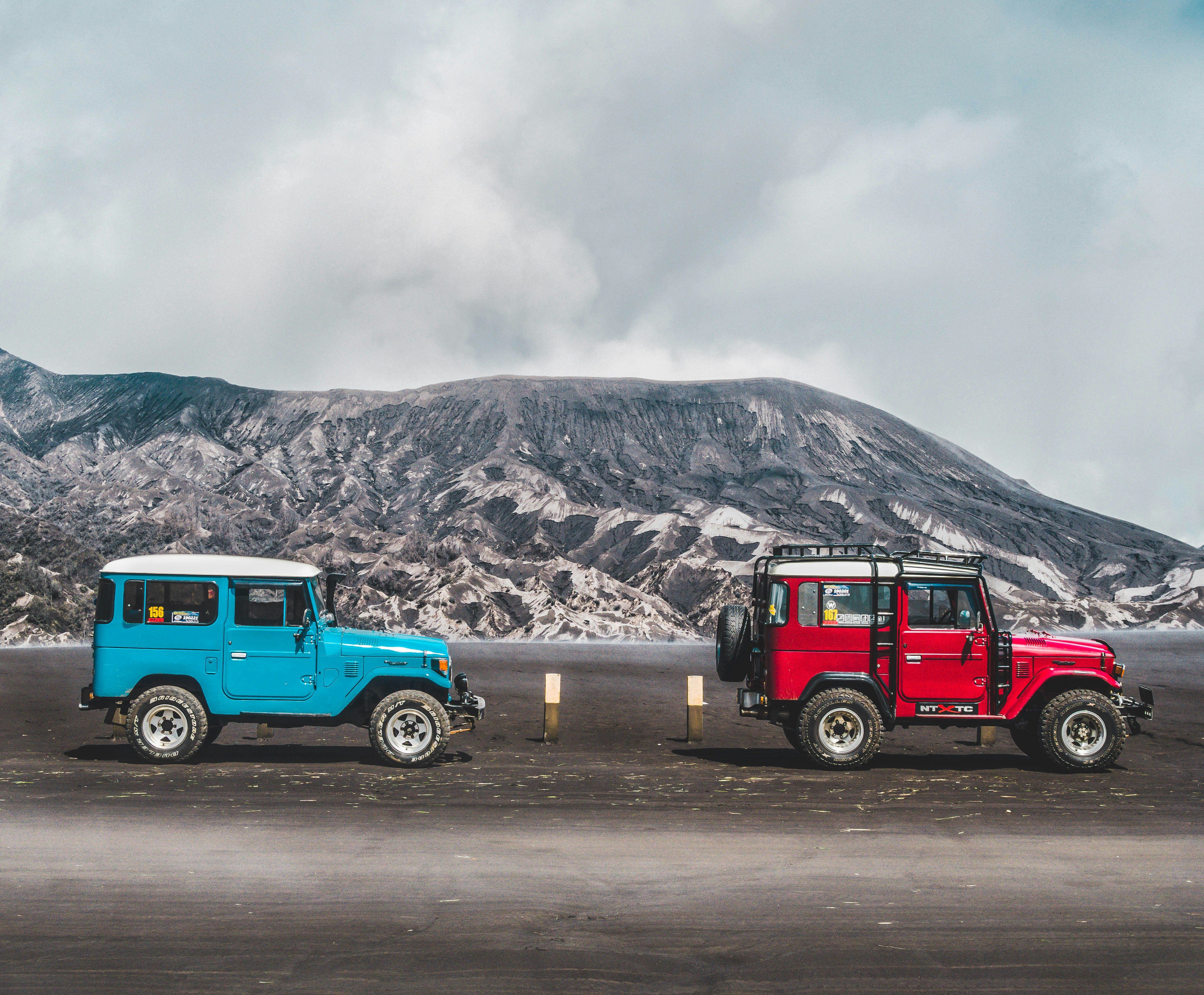 Two vintage off-road vehicles, one blue and one red, parked on a volcanic landscape with mountains in the background.