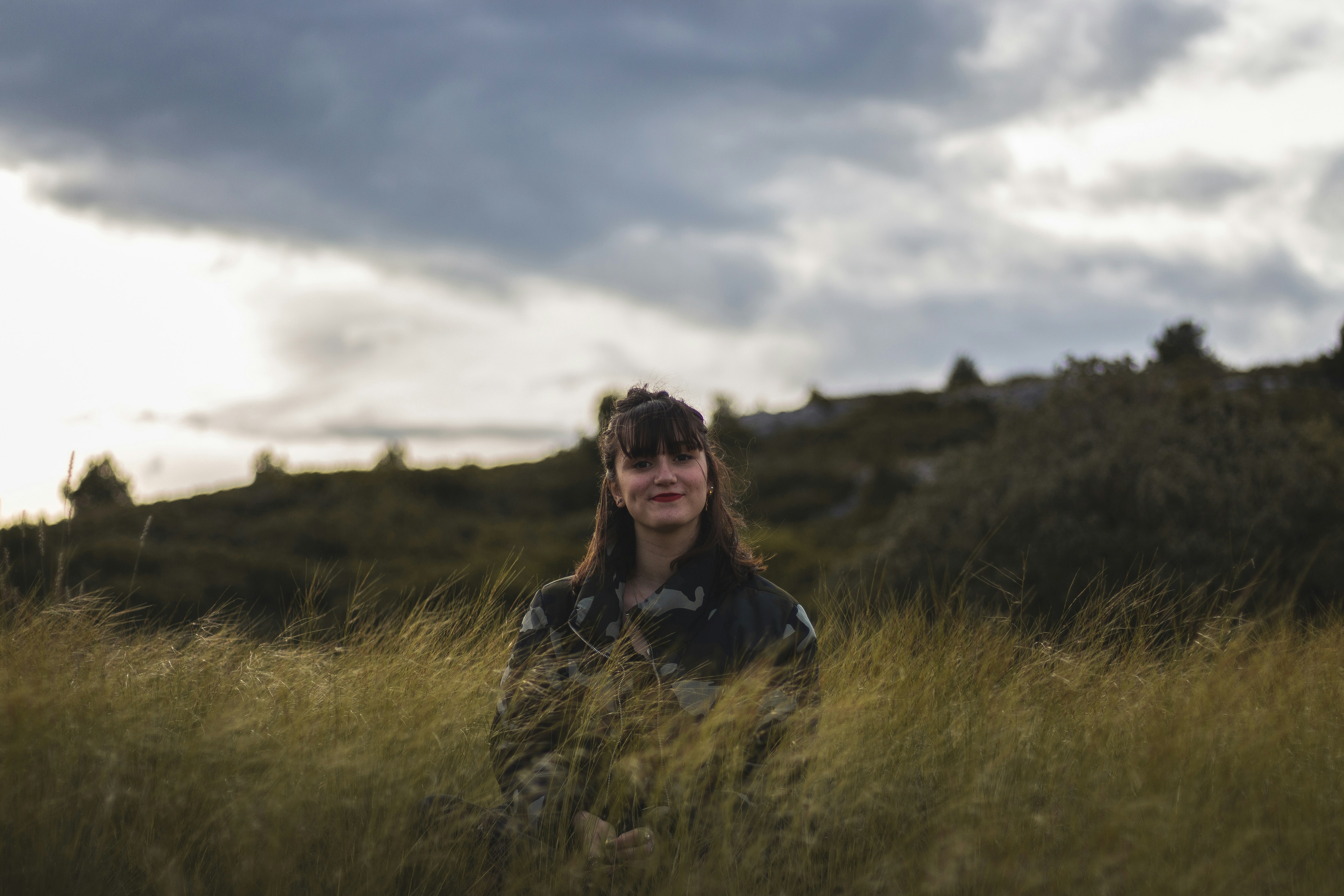 Person in a dark jacket sitting in a field of tall grass under a cloudy sky.