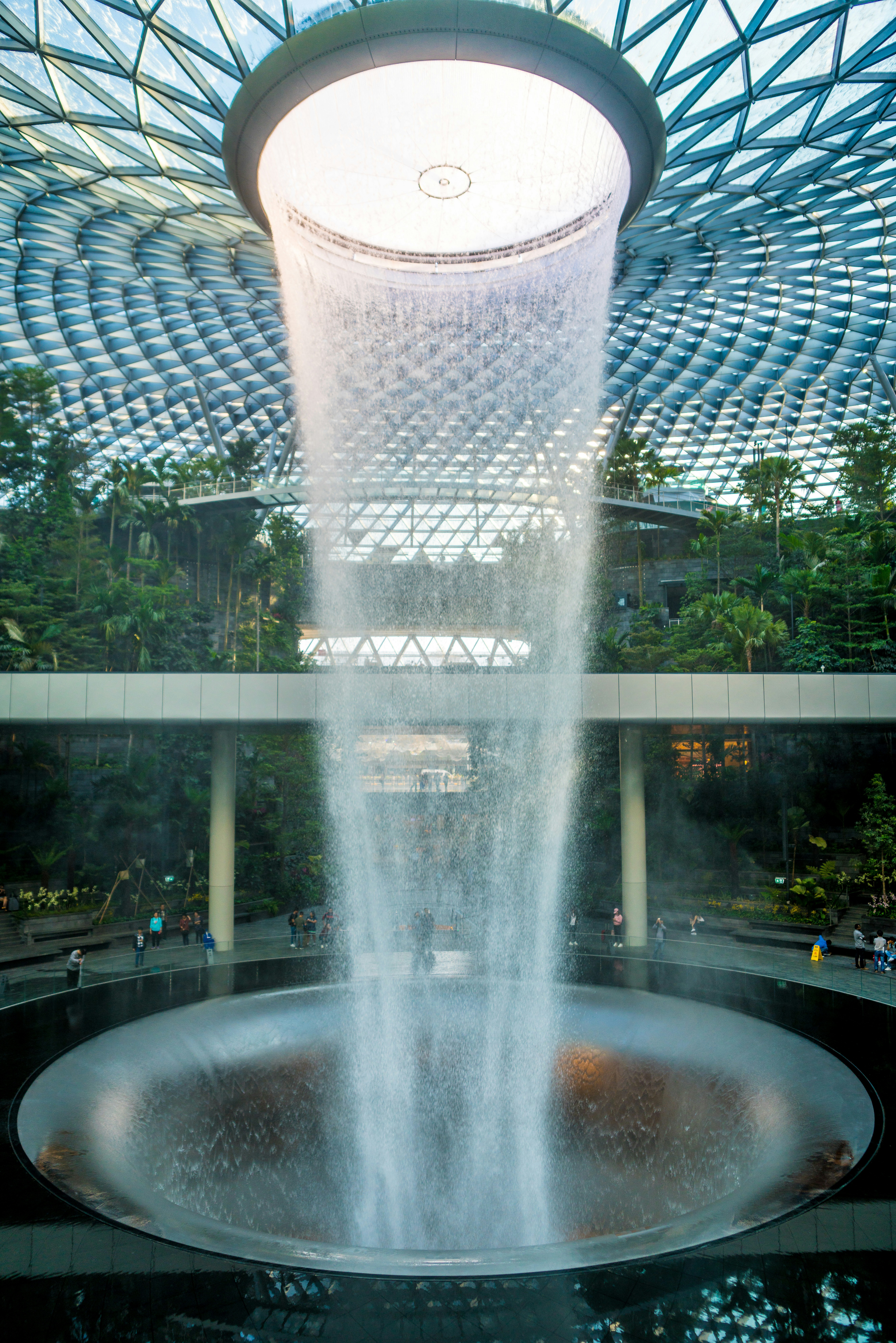A stunning indoor waterfall cascades from a circular skylight, surrounded by lush greenery and a reflective pool below.