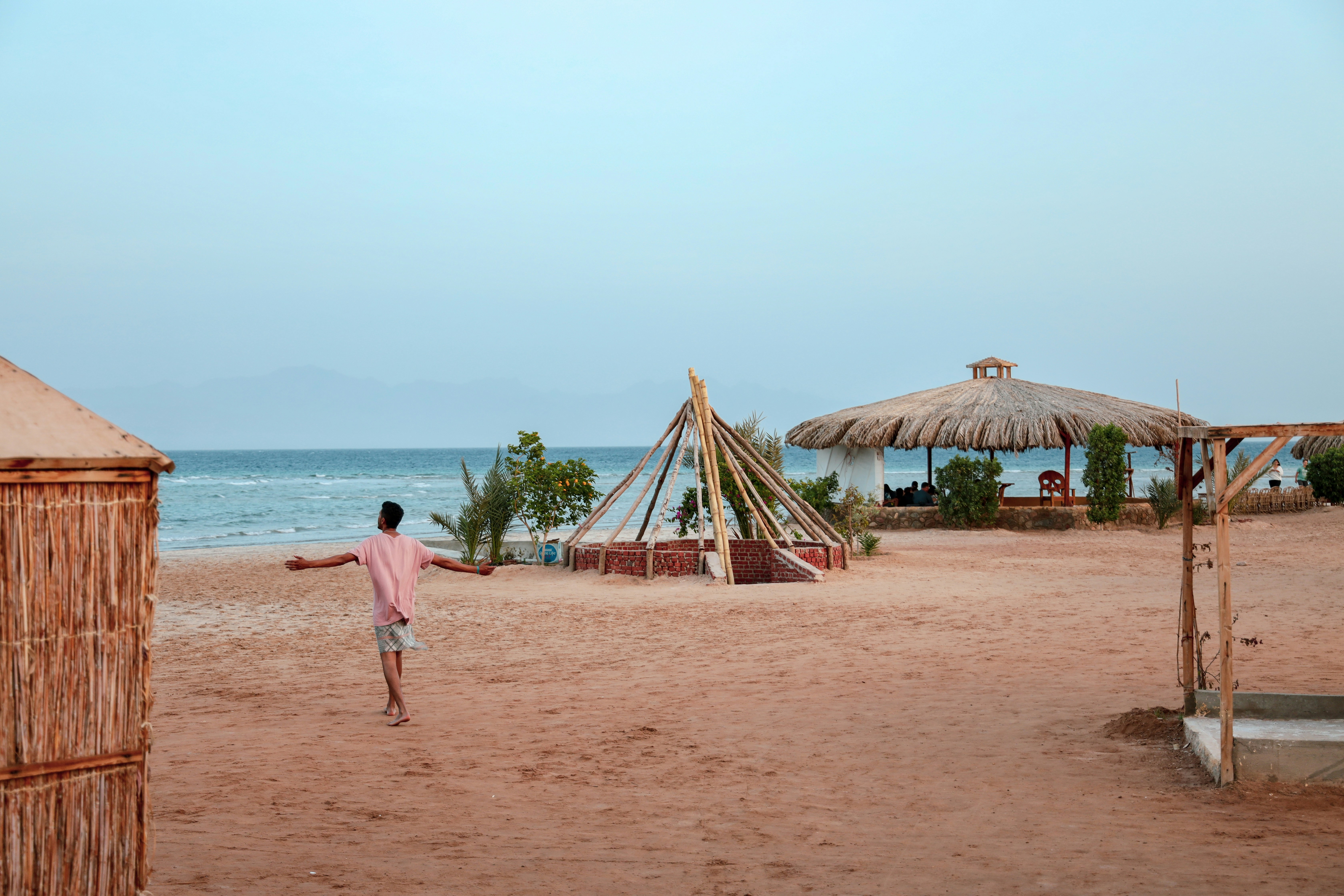 A man strolls along a sandy beach, with a thatched gazebo and a unique structure in the background, embodying tranquility by the sea.